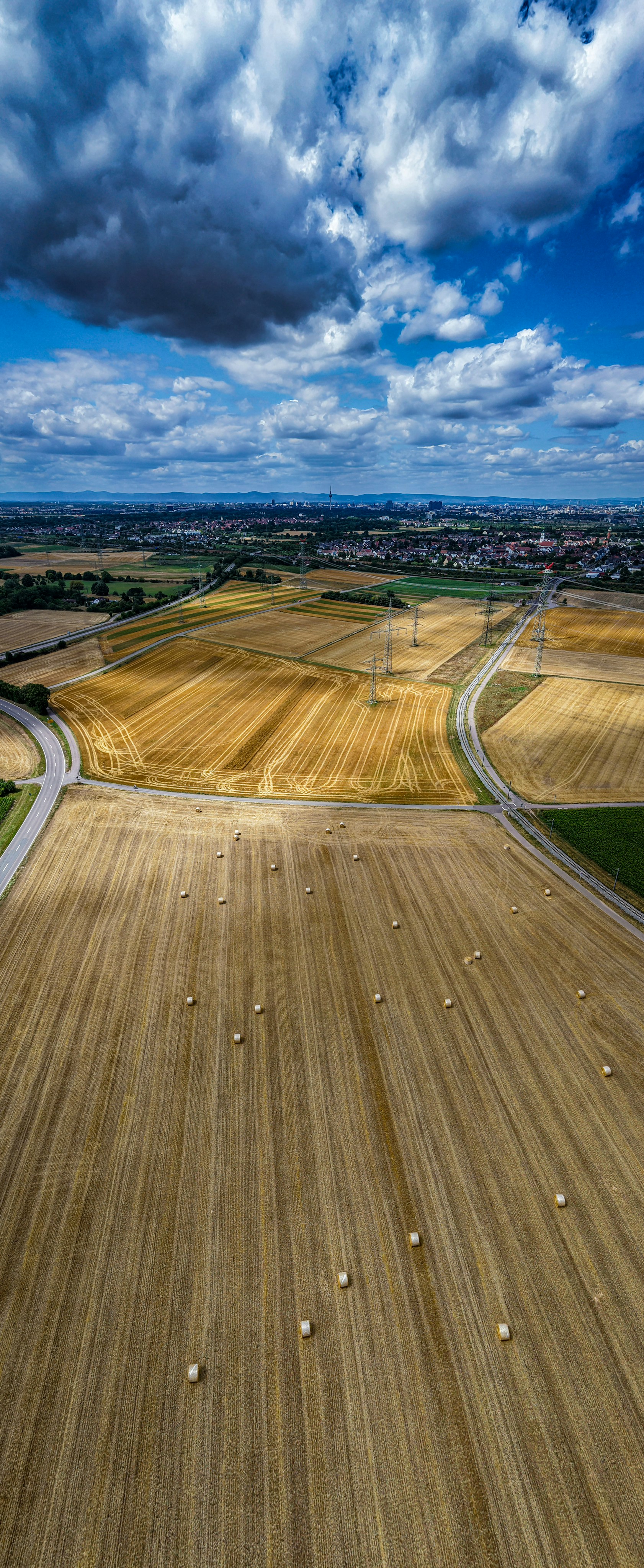 A drone capture of expansive hay fields with round bales, network of roads and distant town under a dramatic cloudscape.