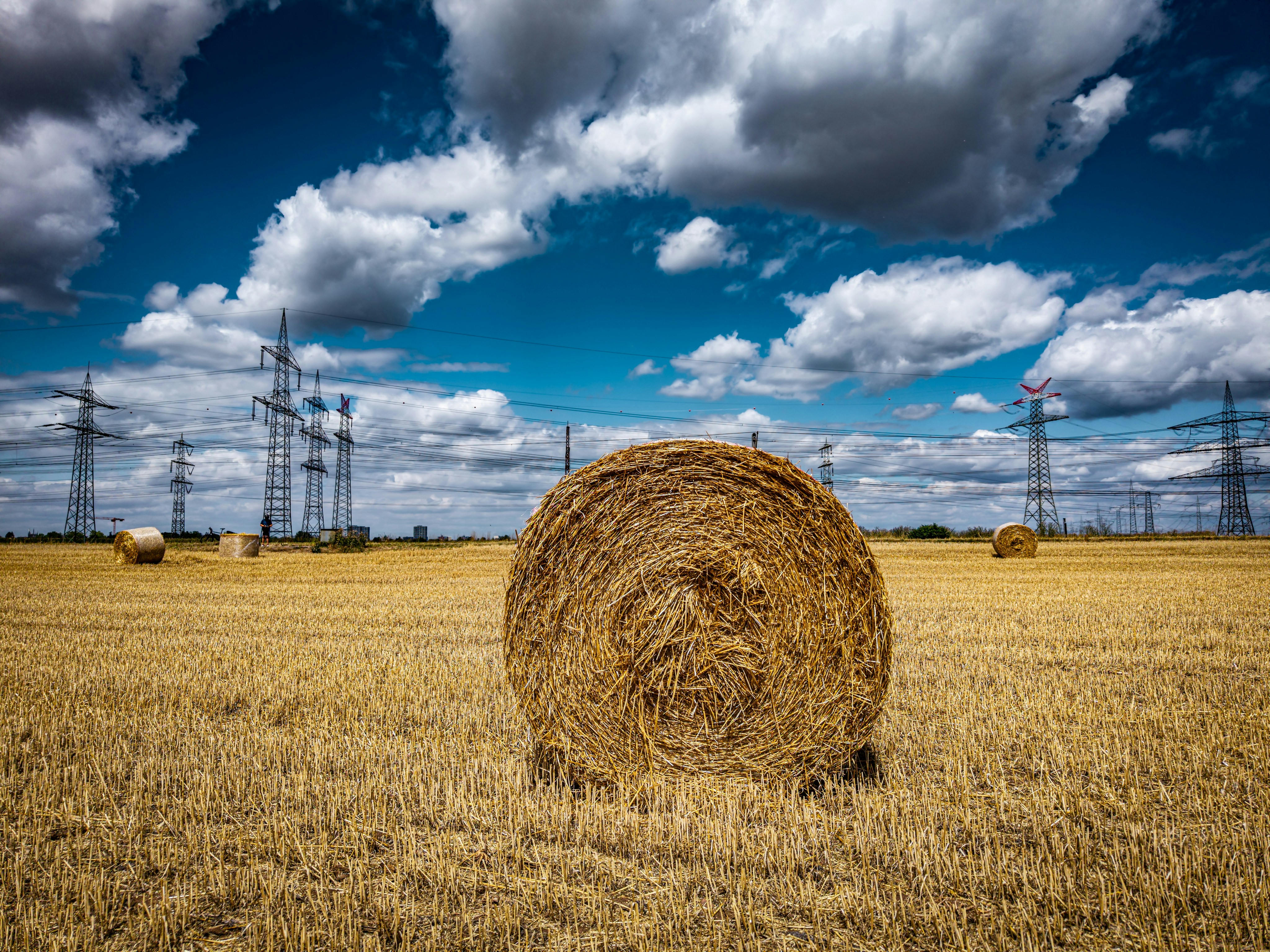 Golden field with a large central hay bale under a dramatic blue sky with clouds; a rural landscape featuring additional bales and distant power lines. This is a photography capture of a farm scene.
