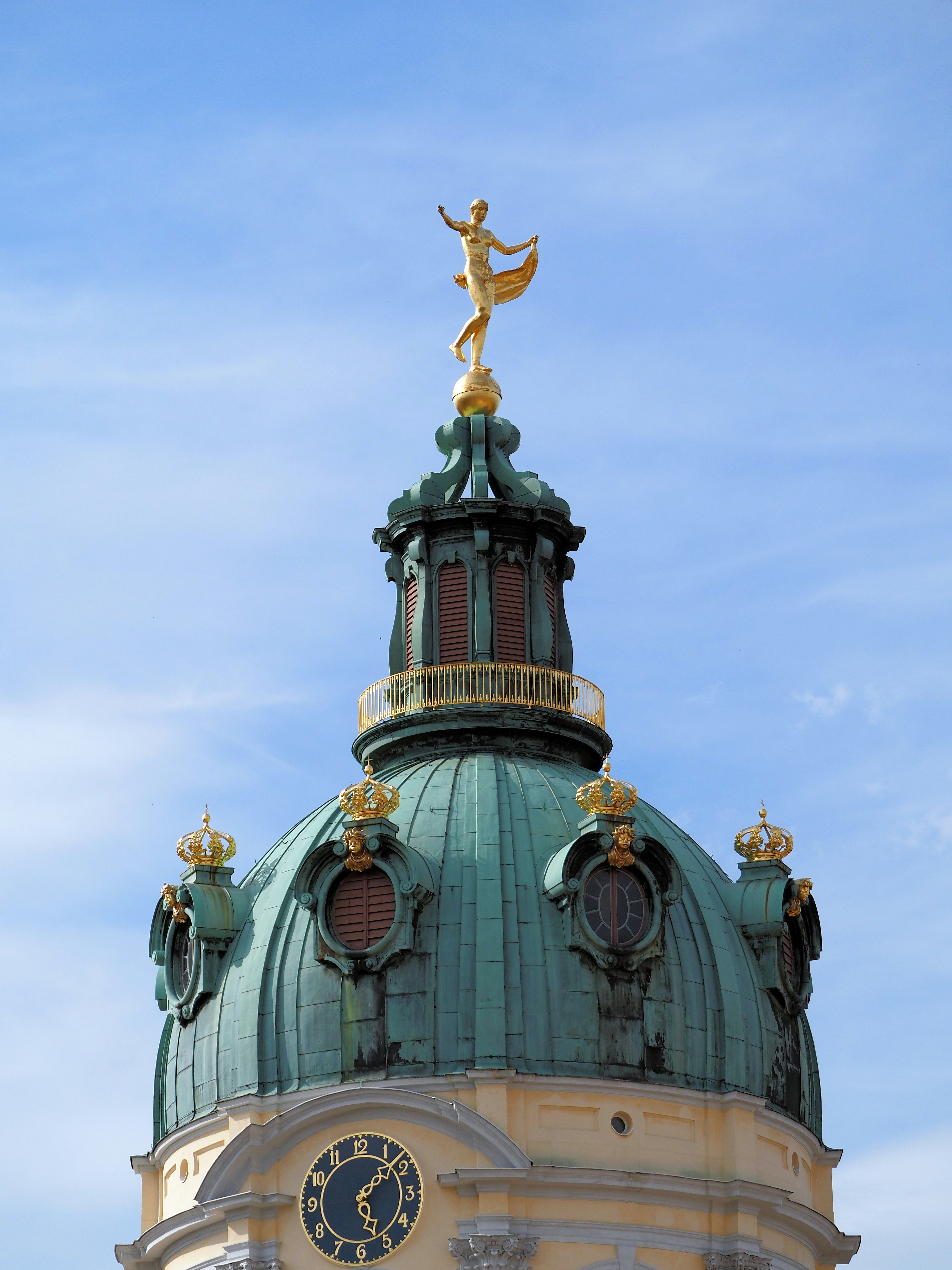 Architectural detail of a domed tower with a gilded statue on top and a clock near the base; the image emphasizes the ornate green roof and decorative gold accents.