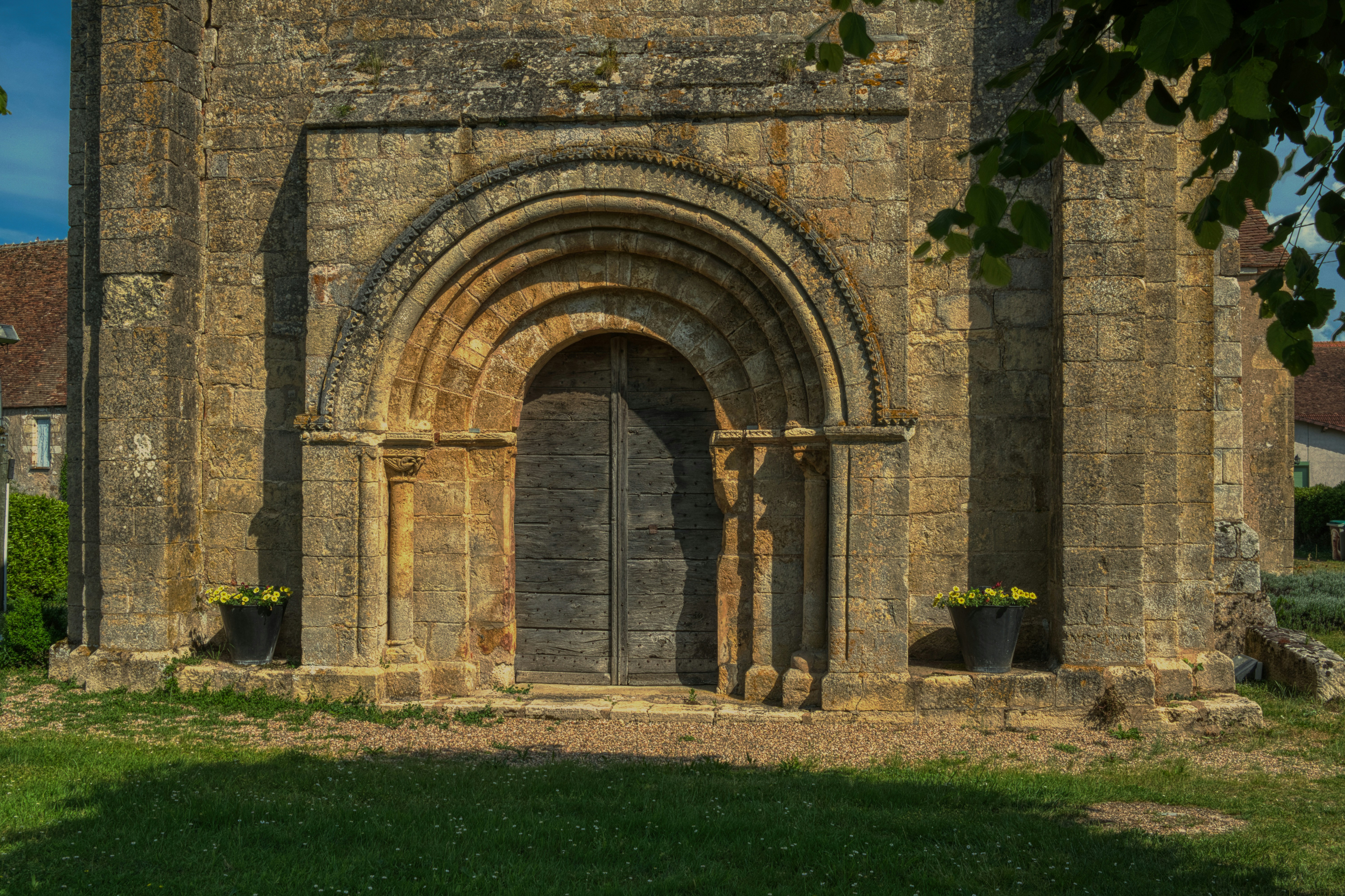 Weathered stone archway with carved concentric arches and heavy wooden doors; two potted plants flank the entrance, set in a sunlit churchyard/Courtyard.