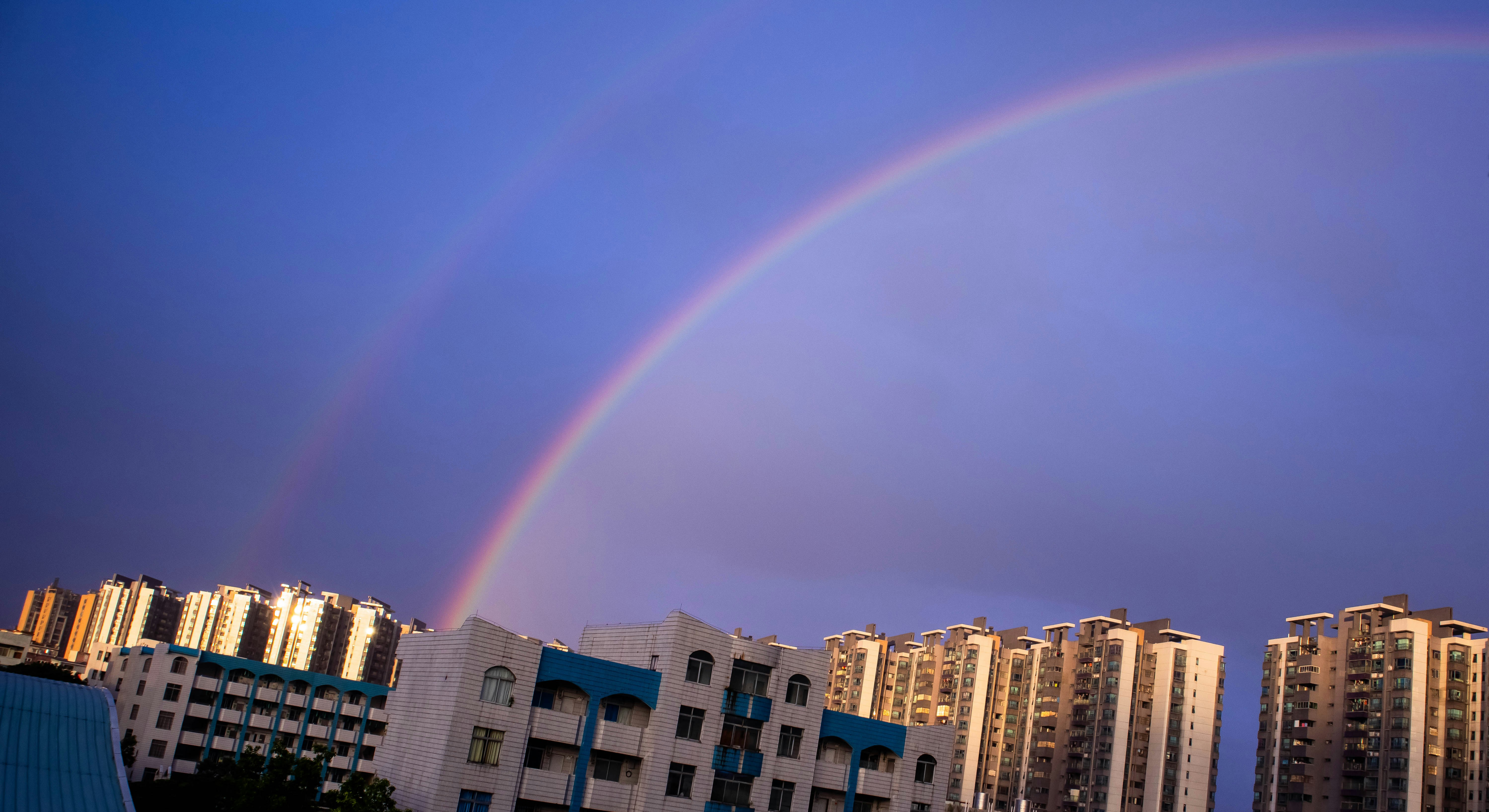 Double rainbow arcs over a row of sunlit apartment buildings, capturing urban skyline beneath a dramatic purple sky.