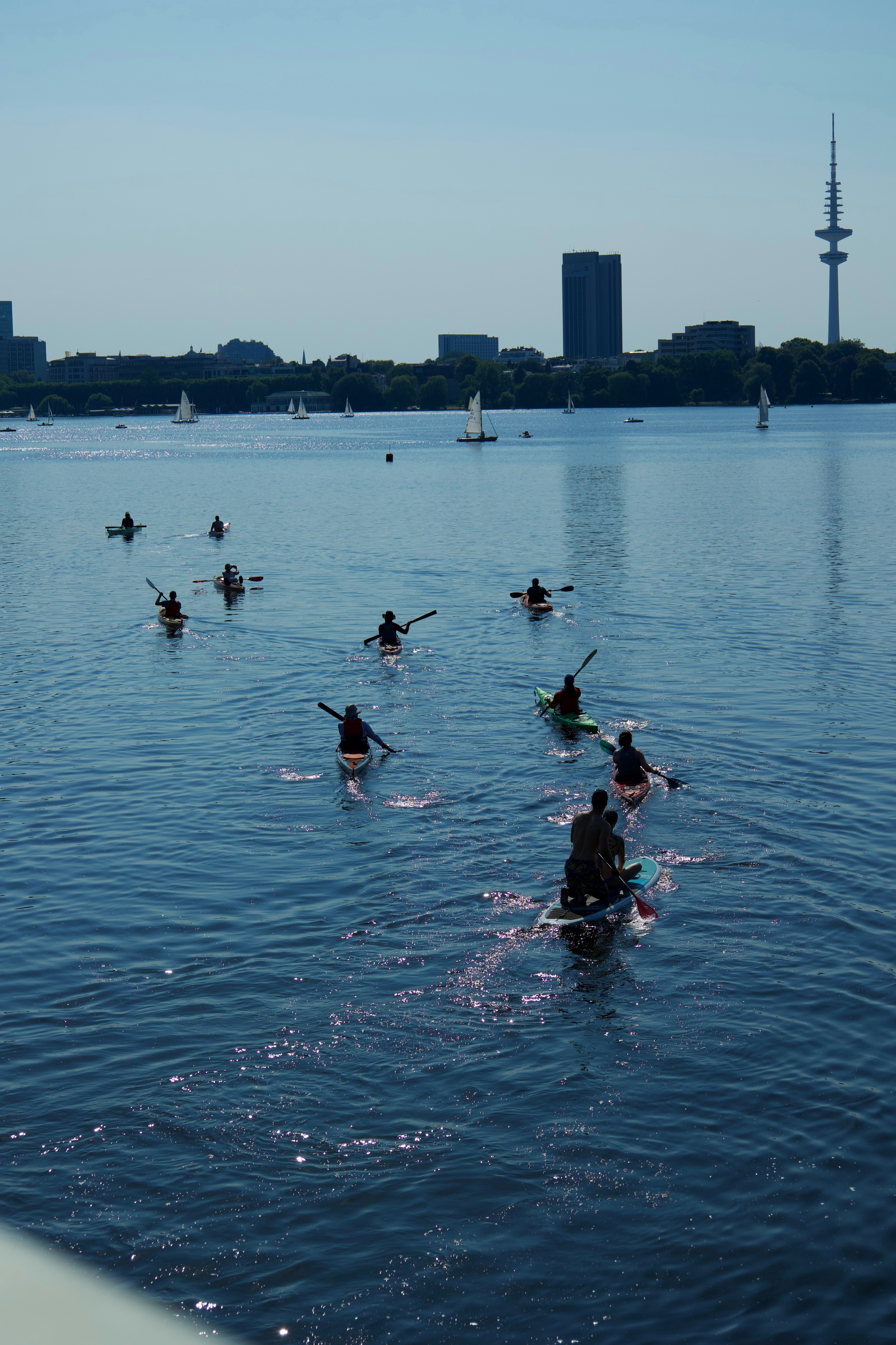 Group of kayakers and small boats moving across a calm lake with a city skyline and tall communications tower in the background; the scene captures active water sport against an urban horizon.