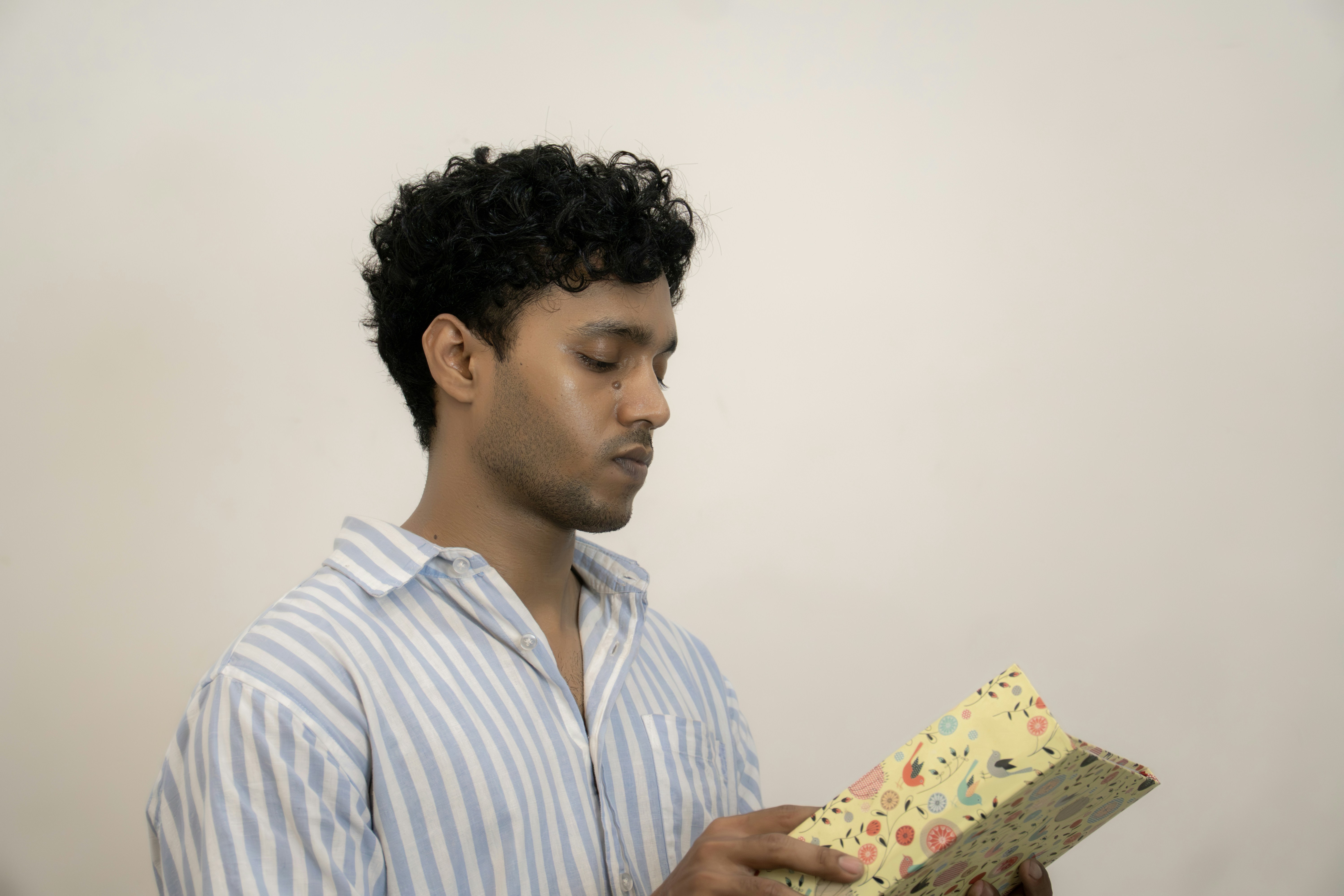 A young man with curly hair wearing a blue and white striped shirt reads a colorful floral notebook while standing against a clean beige background. The image conveys focus, learning, education, creativity, and quiet reflection. Ideal for concepts related to studying, journaling, reading, productivity, lifestyle, self-improvement, and modern youth.