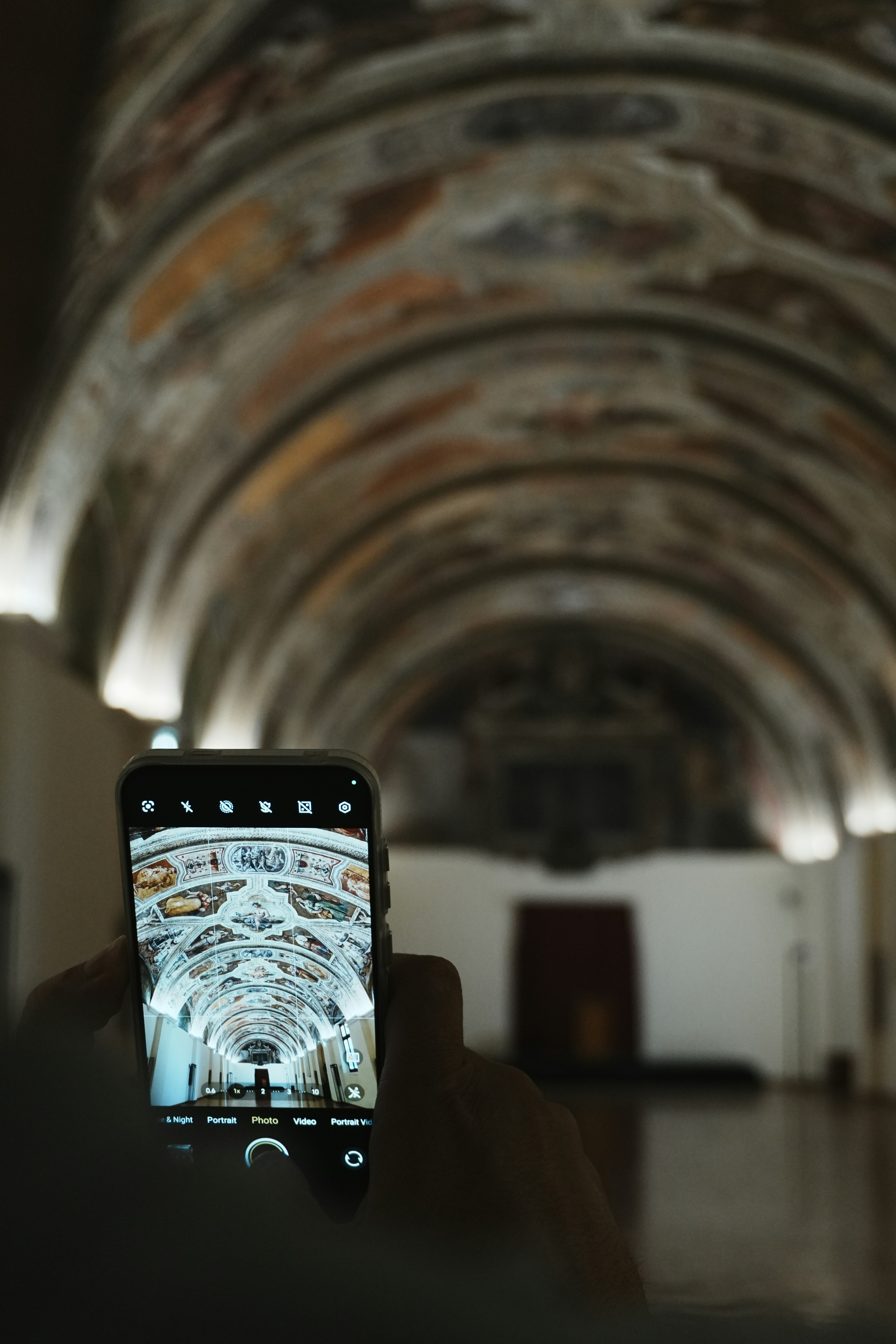 A handheld smartphone frames an arched, ornate ceiling inside a dim interior, focusing on the device screen showing the shot. The scene emphasizes architectural detail and the act of photographing the space.