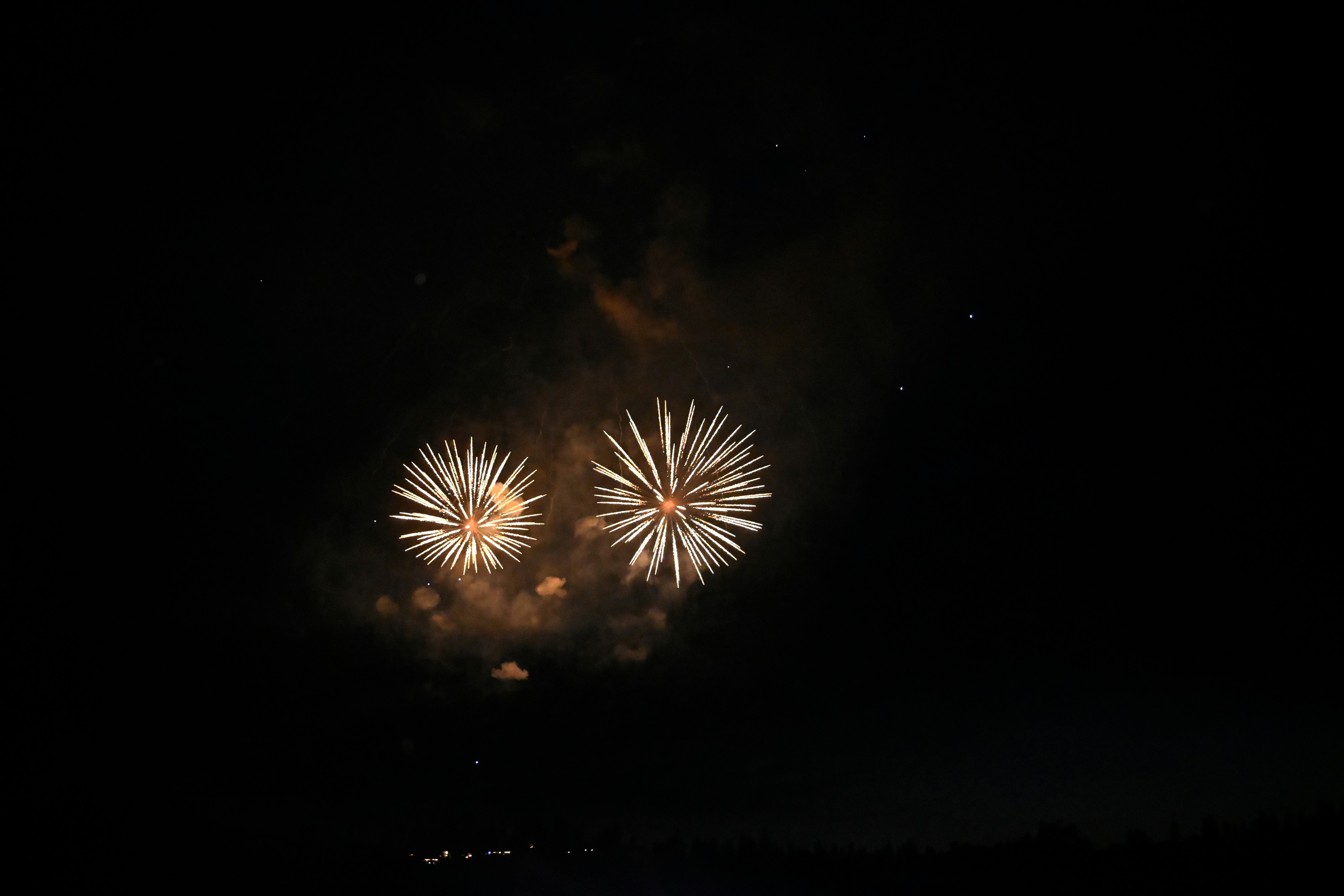 Dark night sky with two bright fireworks explodings above a distant horizon; foreground shows subtle smoke and silhouettes, capturing a celebratory moment.