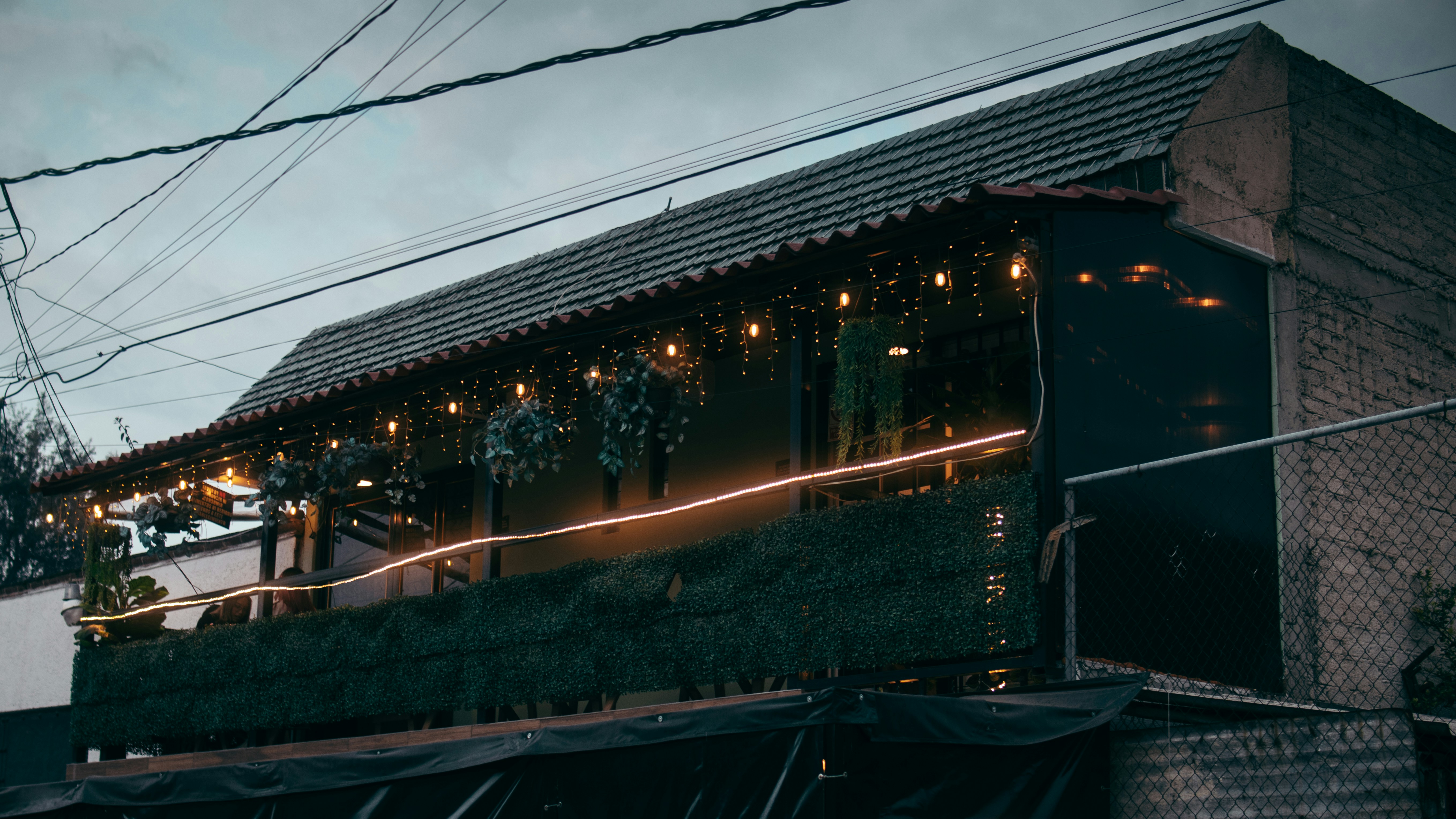 A cozy balcony of a brick building adorned with string lights and greenery, capturing a quiet urban moment at dusk.