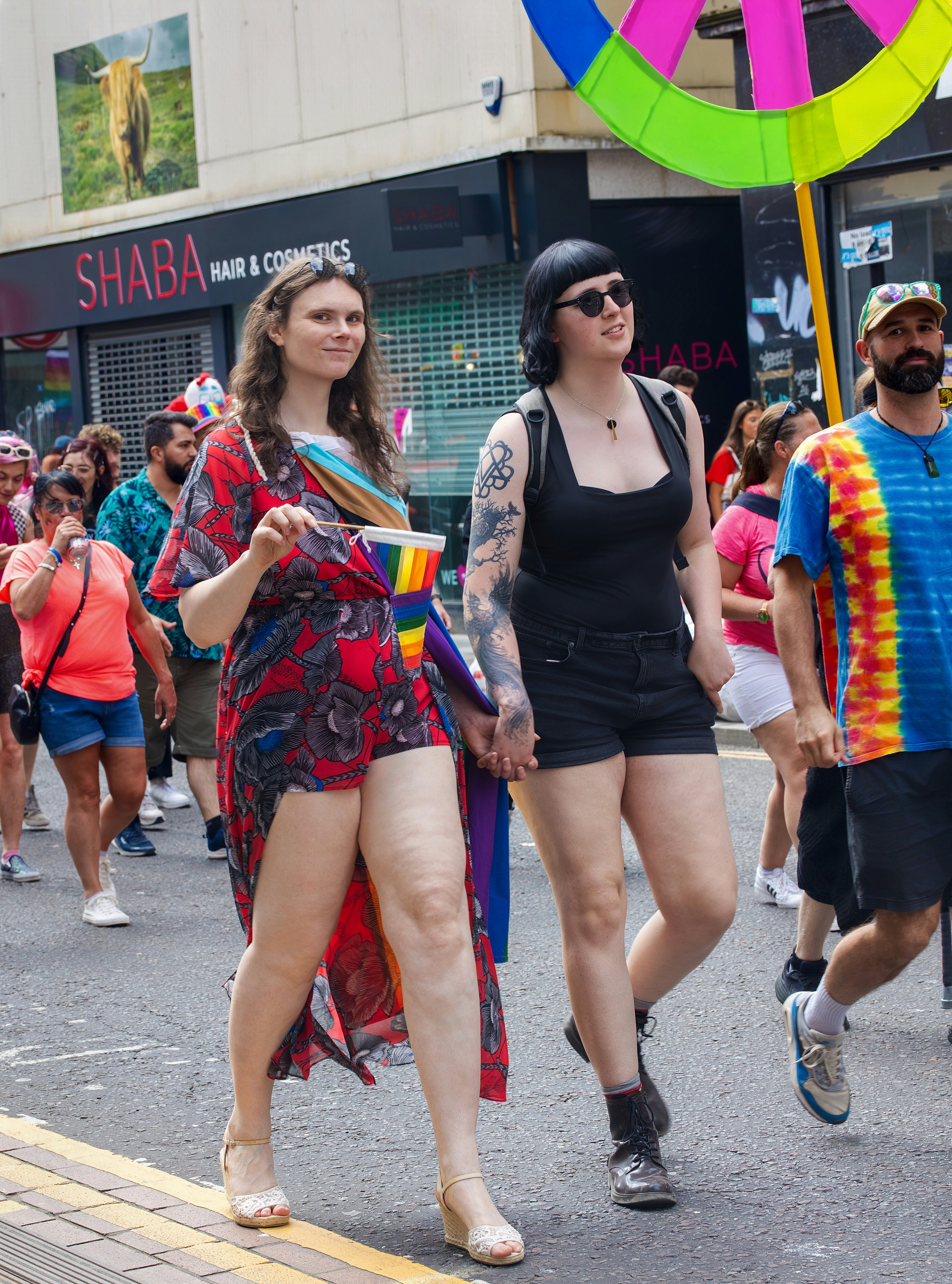 Two festival-goers stride forward hand in hand beneath a rainbow roundel