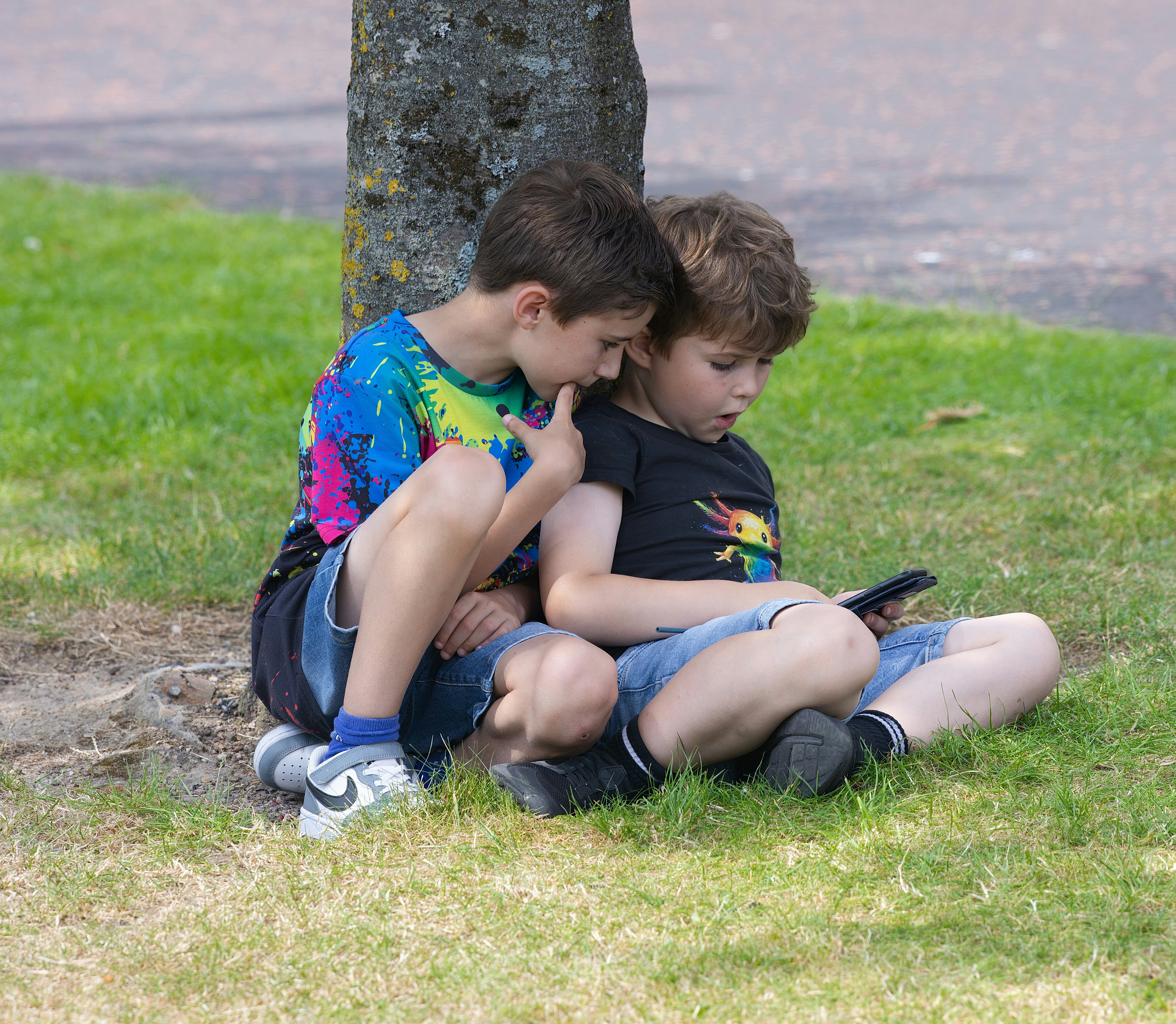Two young boys sit on grass against a tree, absorbed in a handheld device, while wearing casual summer clothes in a park-like setting.