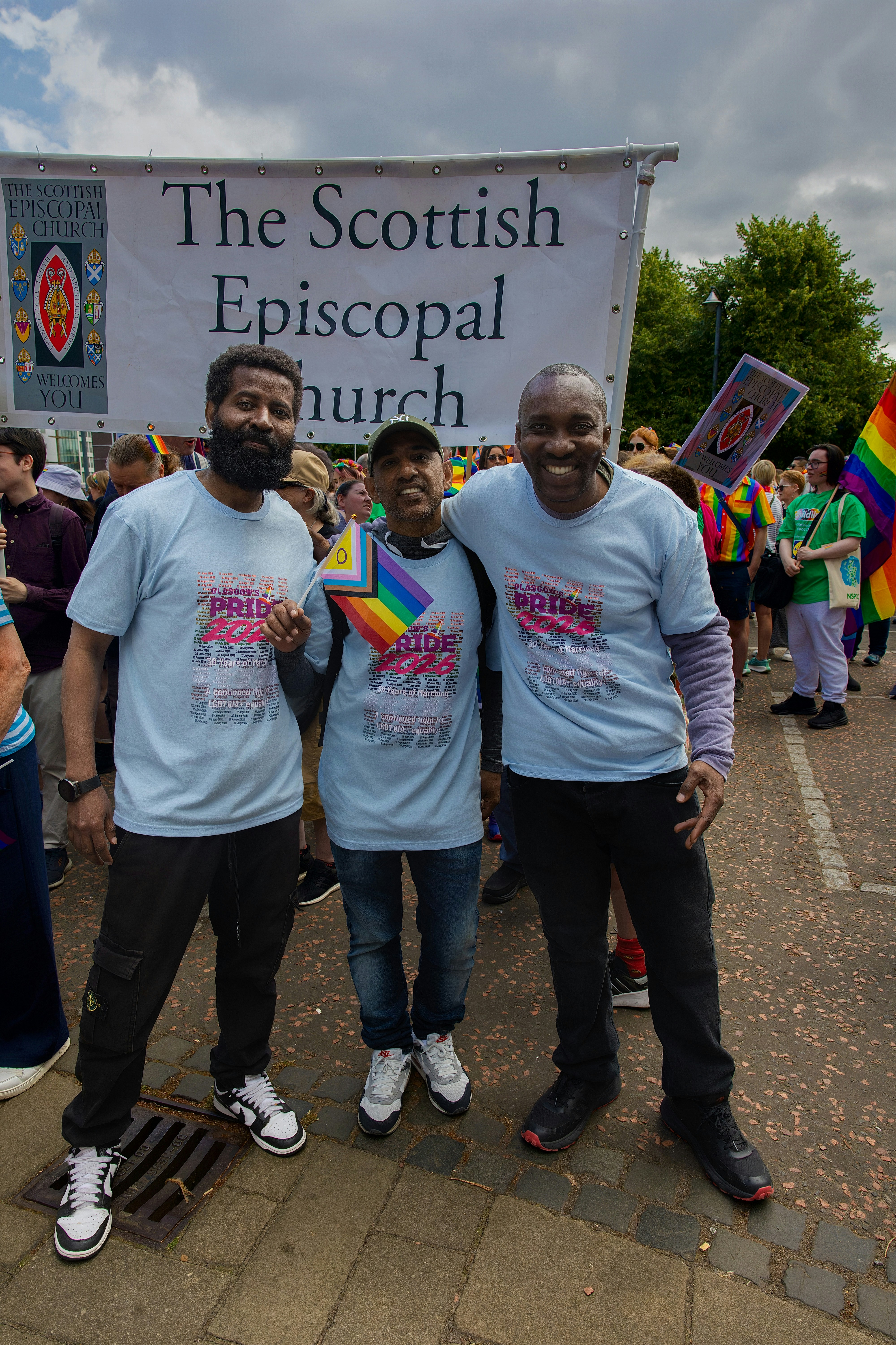 Three Friends in Pride Parade T-Shirts Pose Beneath The Scottish Episcopal Church Banner