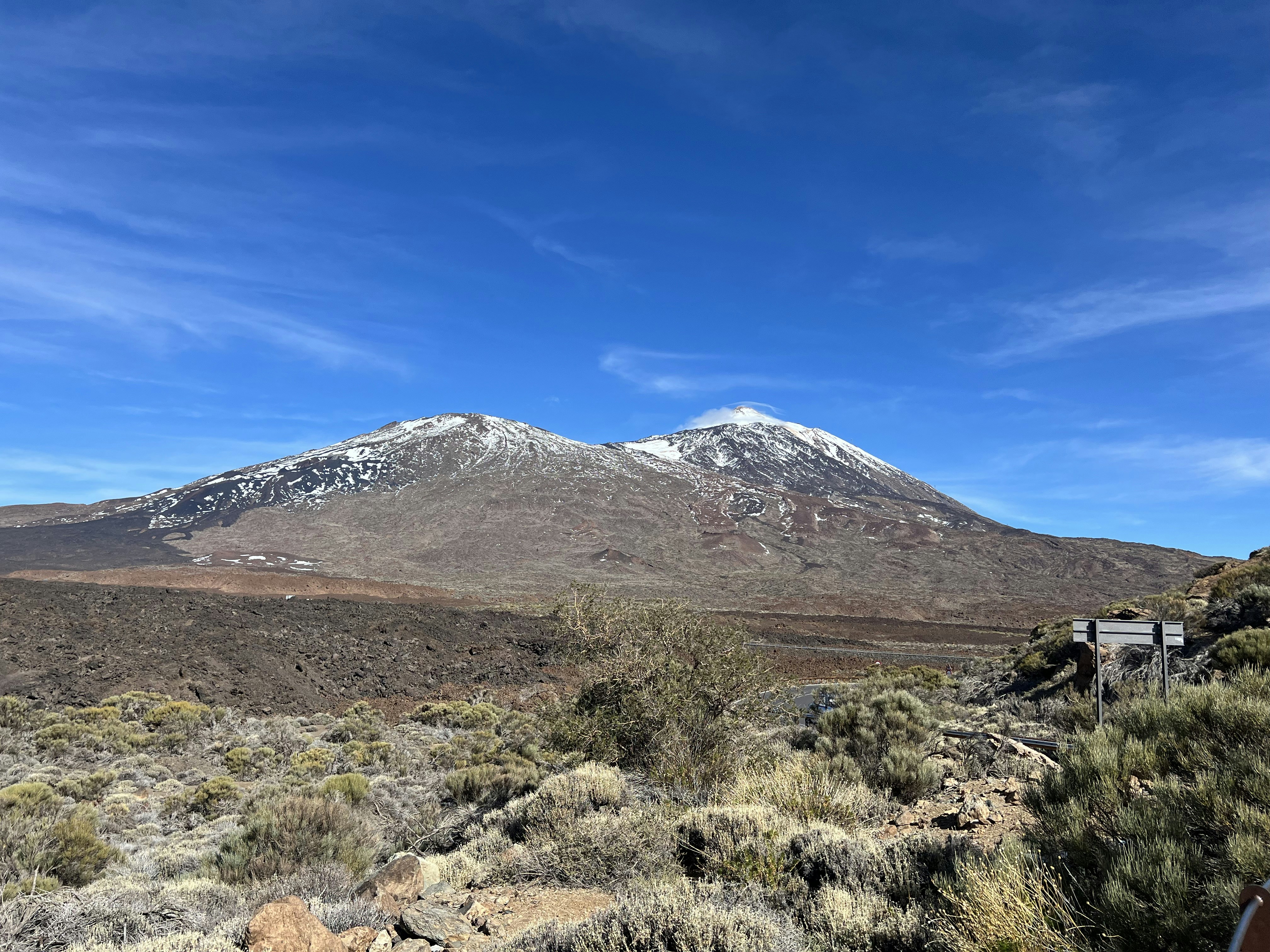 Desert scrub foreground with distant snow-tipped mountains under a bright blue sky; rugged terrain and a small structure hint at human presence.