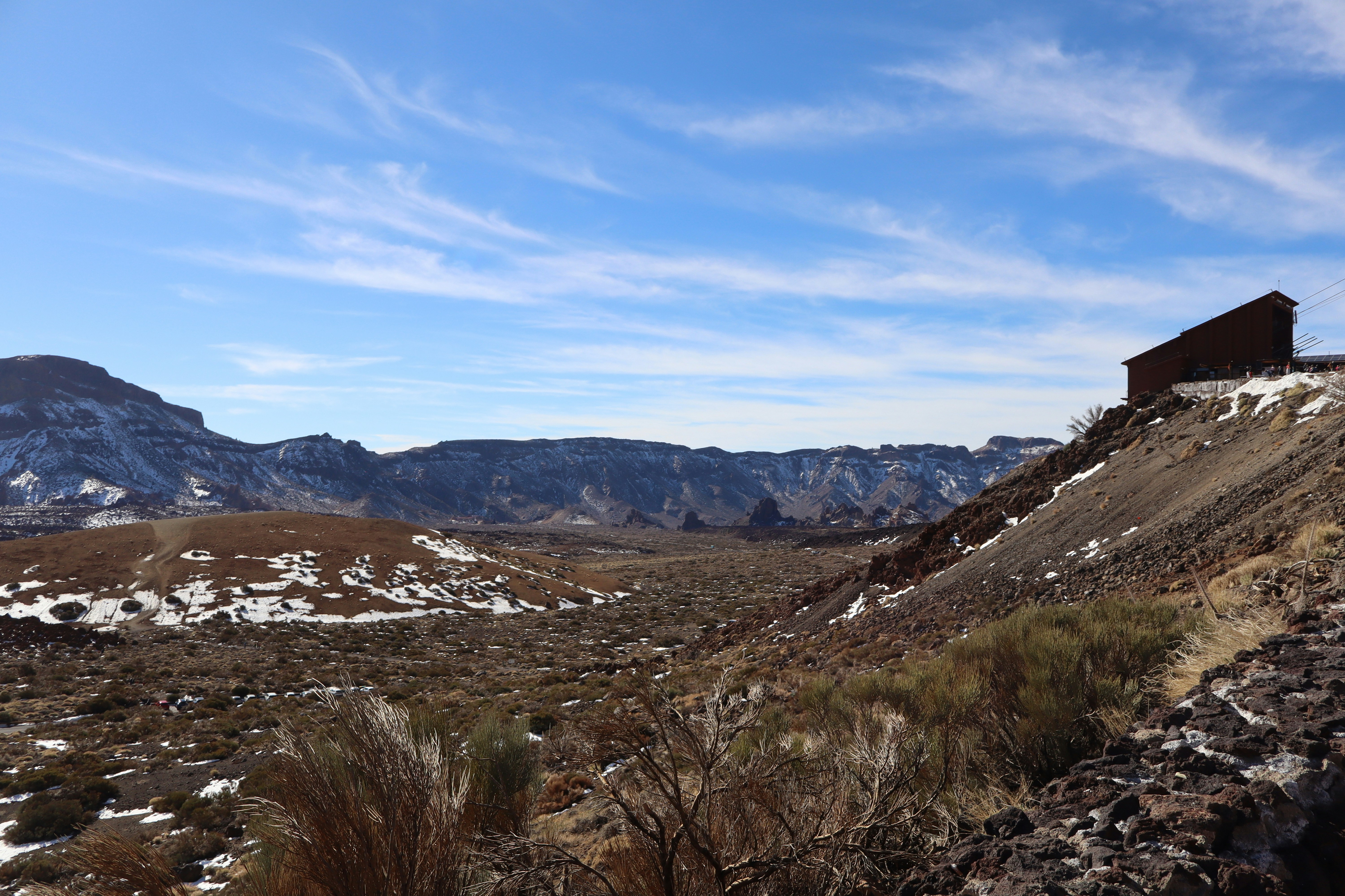 Sunlit desert valley with scrubby foreground leading to rugged hills and a small building perched on a rocky slope; distant mesas under a bright blue sky.