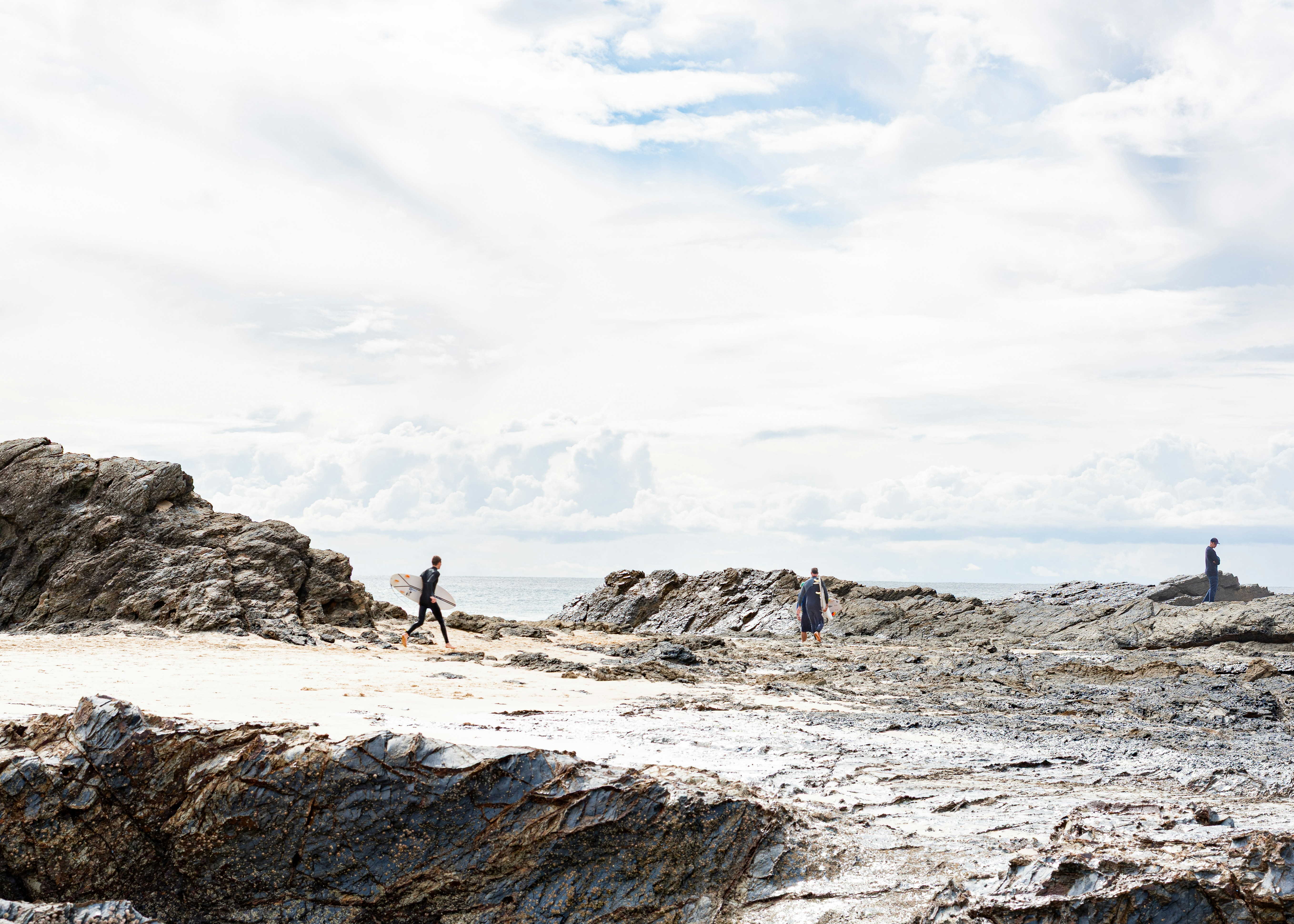 Three figures traverse a rocky beach with rugged outcrops under a bright, airy sky; the scene emphasizes scale and rugged coastline. This is a photographic capture of a coastal landscape.