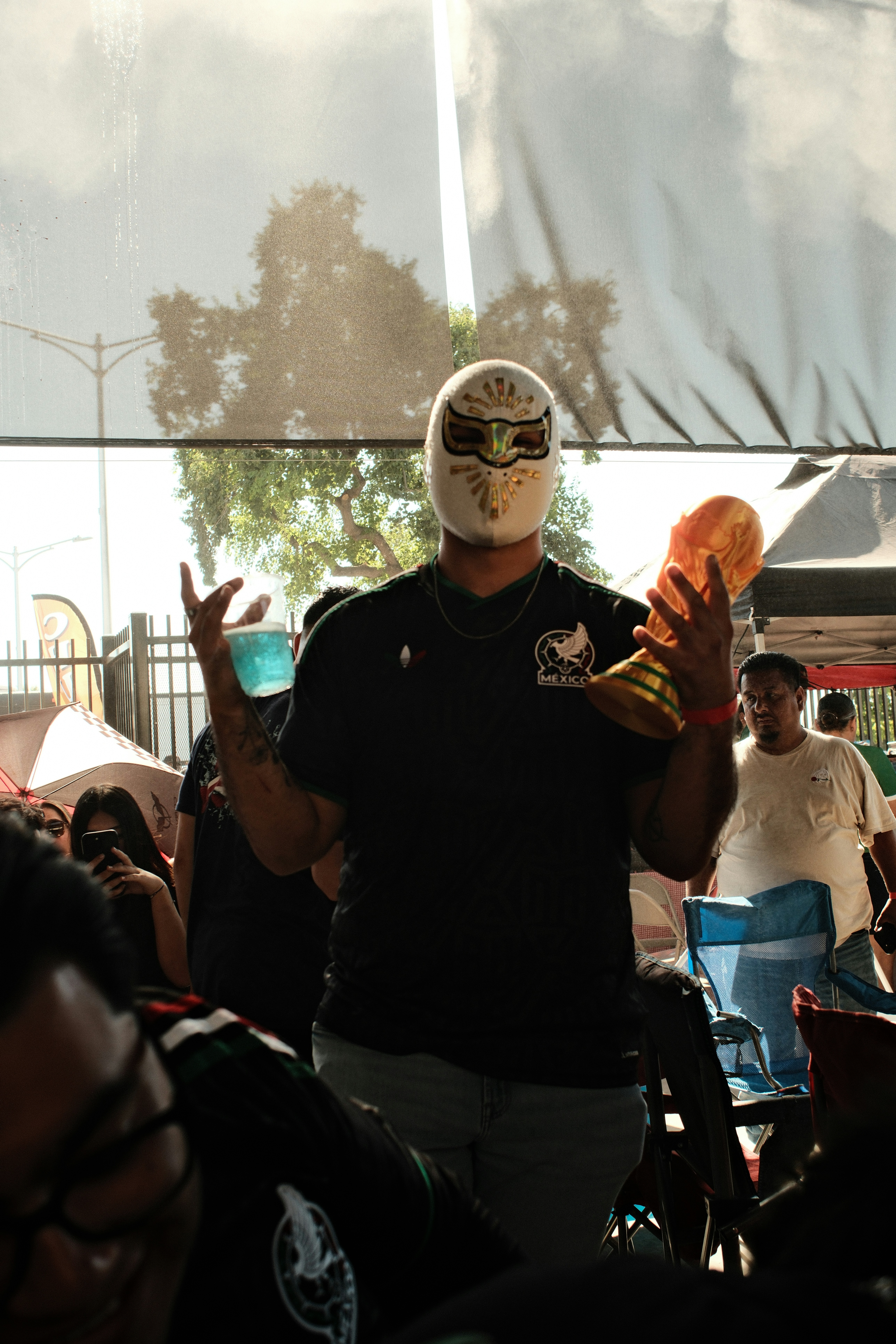 A masked performer stands center-stage under a tent, holding a glowing cup in one hand and a carved object in the other, surrounded by spectators and ambient festival light.