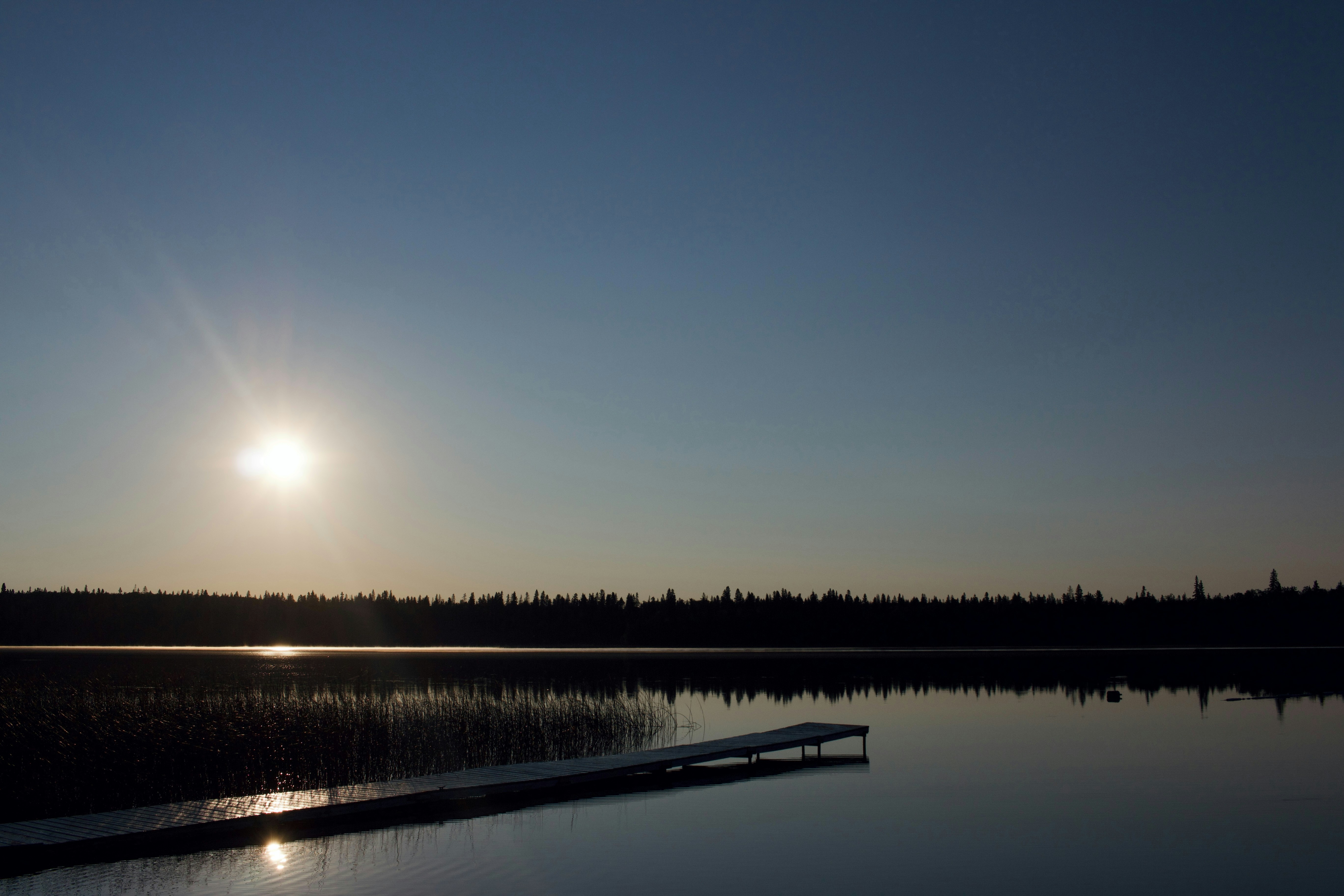 Calm lake scene at sunset with a wooden dock extending into still water; a distant tree line silhouettes the horizon.
