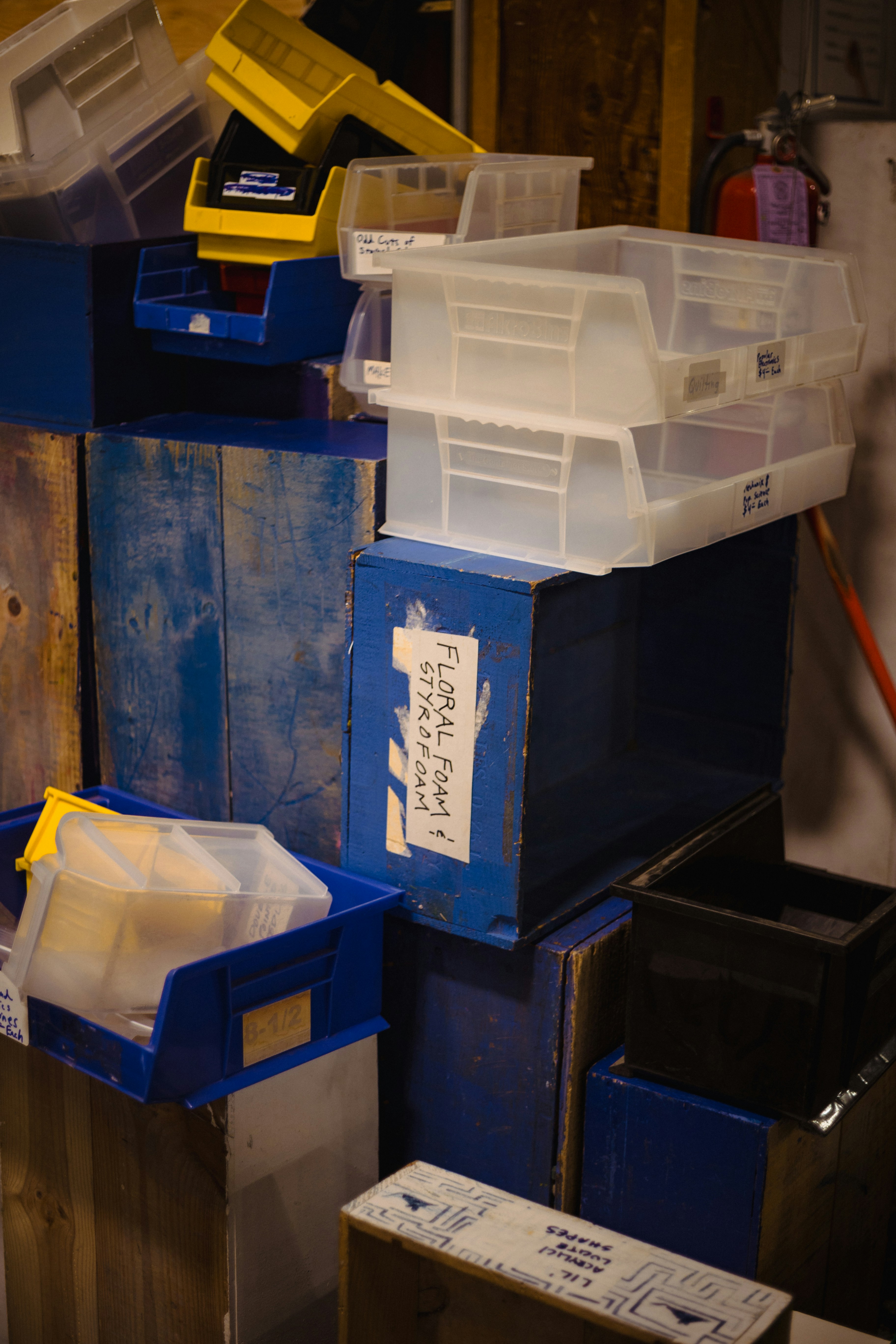 A cluttered storage area with stacked translucent containers and labeled blue crates in a workshop setting. Plastic bins and tool organizers occupy the frame, suggesting a workspace organization scene.