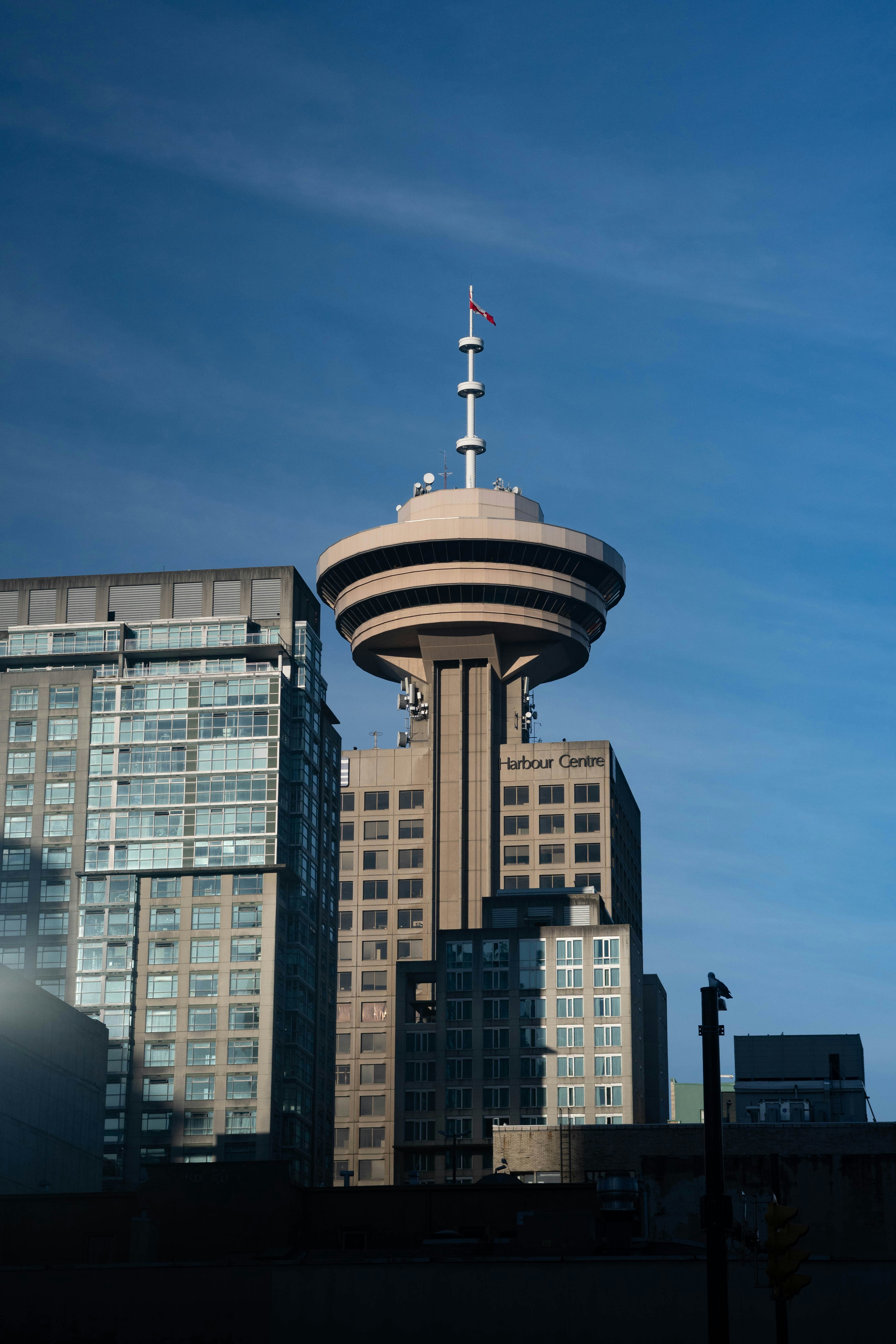 Dramatic urban scene with a distinctive circular observation deck rising above modern glass buildings against a clear blue sky. The composition emphasizes architectural geometry and vertical scale.