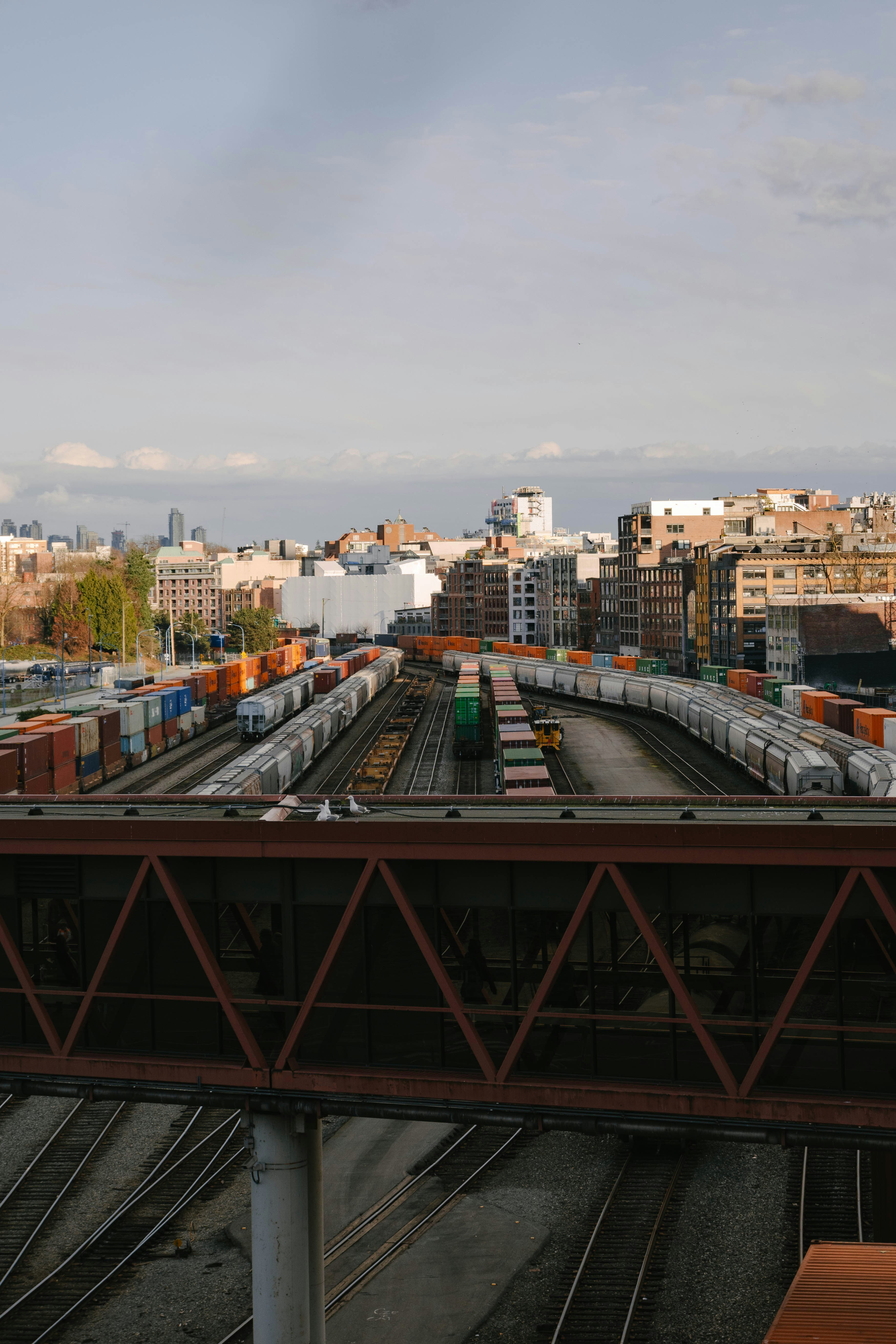 A sweeping view of a freight rail yard with stacked shipping containers, elevated track, and a distant cityscape under a soft evening sky.