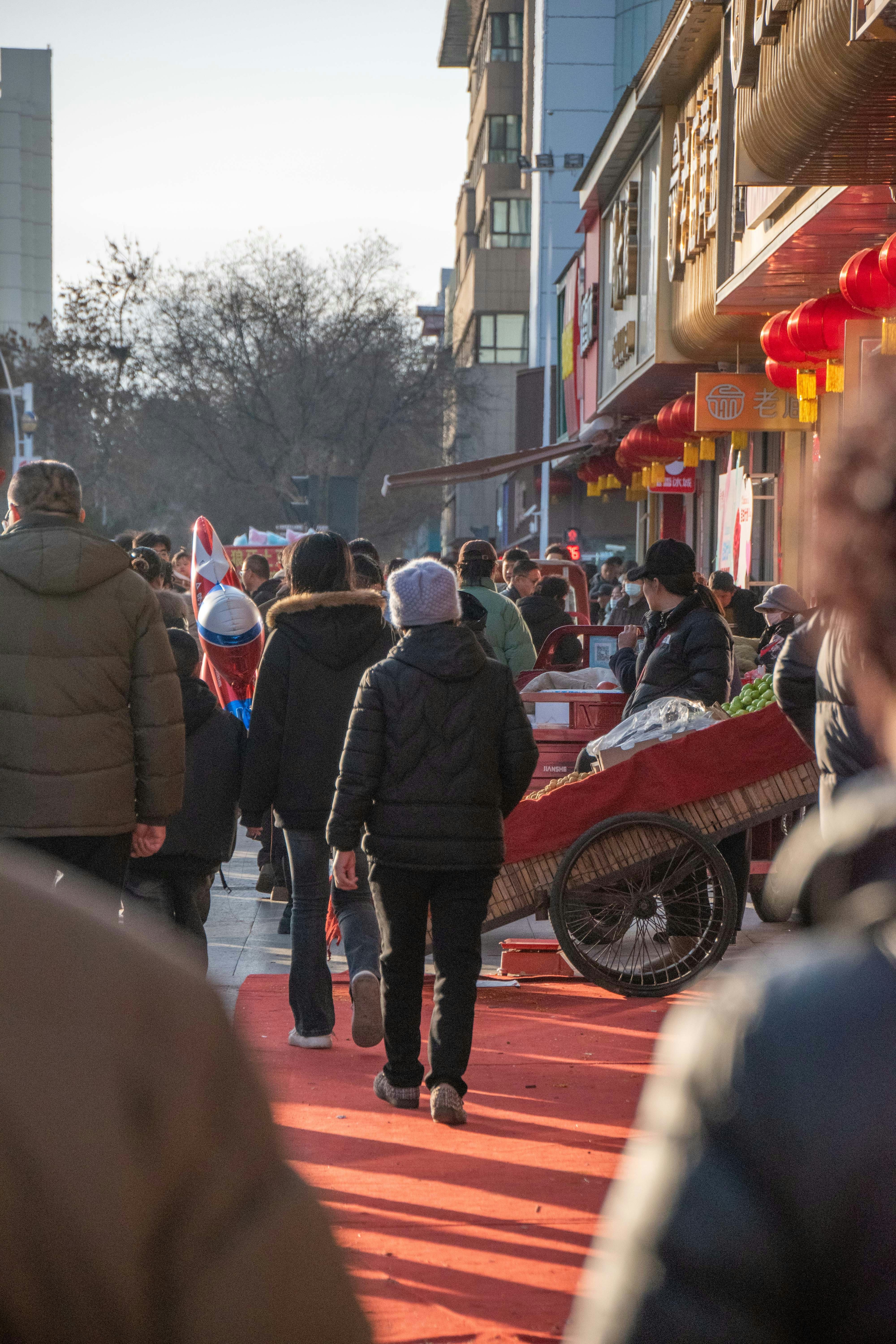 Bustling street scene with pedestrians along a red carpet and lantern-adorned storefronts, capturing daily life with a wheel cart in the foreground. This is a candid photograph of a busy market street.