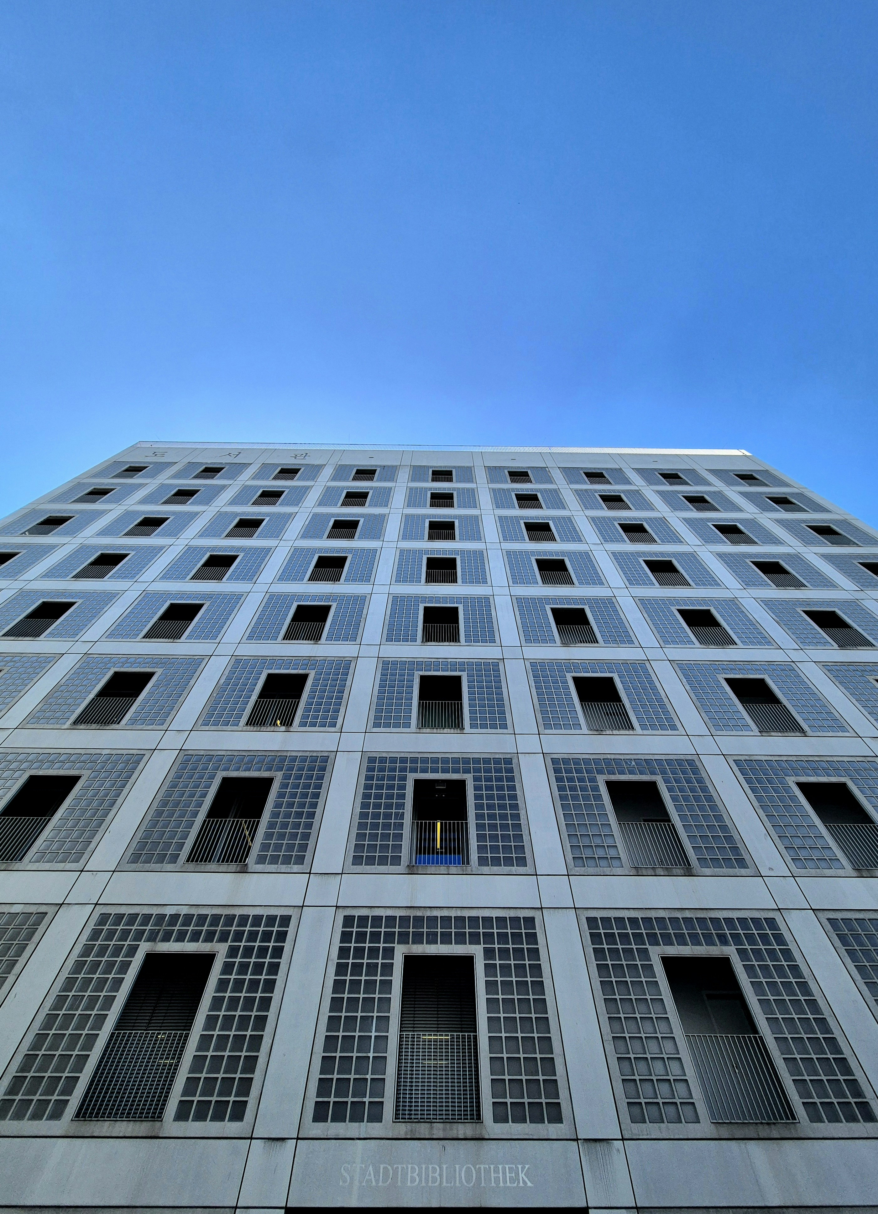 Brutalist facade with repetitive grid windows rises toward a clear blue sky; the photo emphasizes architectural rhythm and texture of the building's panels. The structure is identified as a municipal library.