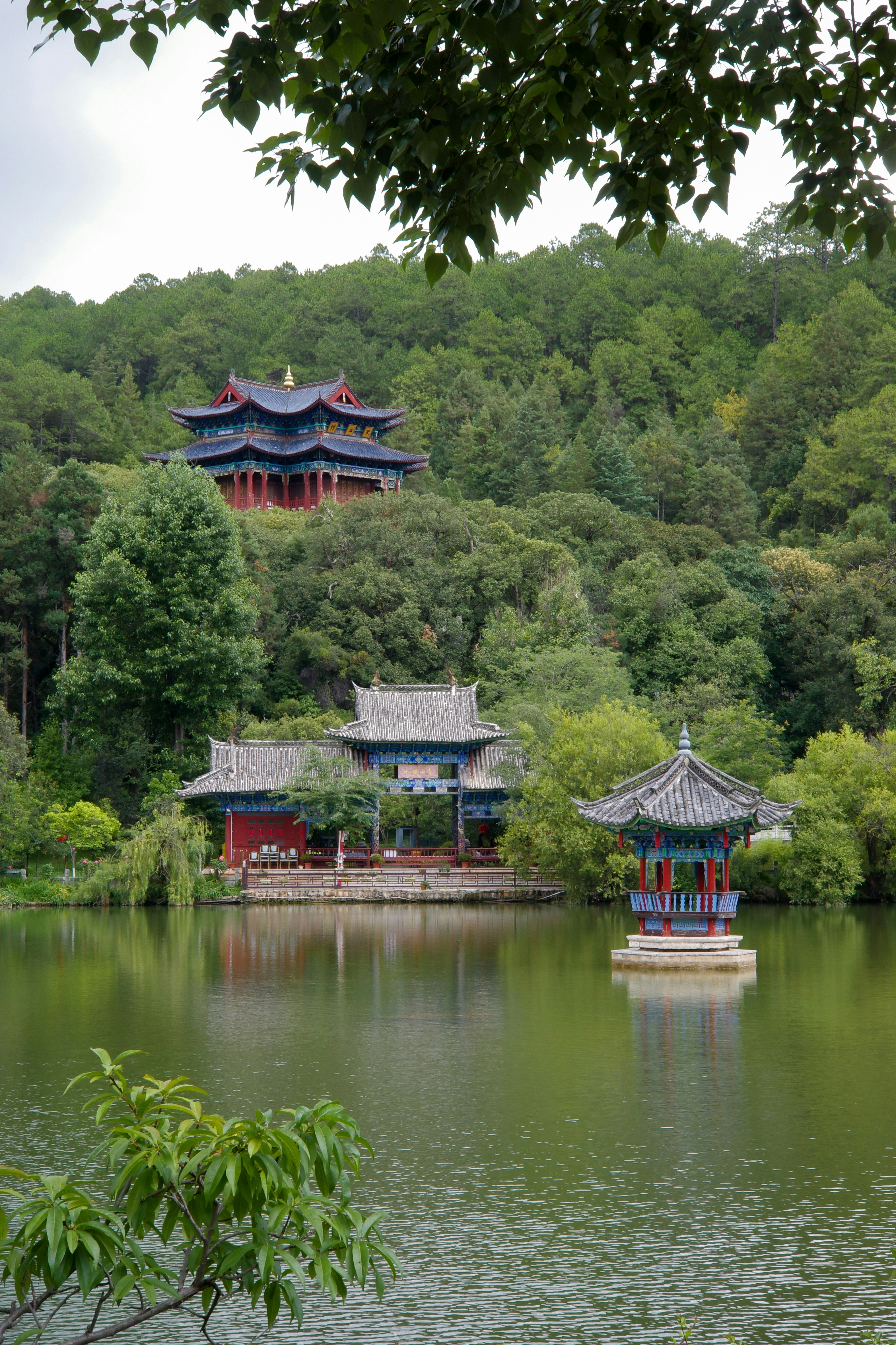 Colorful pavilion rising from the lake, framed by layered greens and distant hillside temples