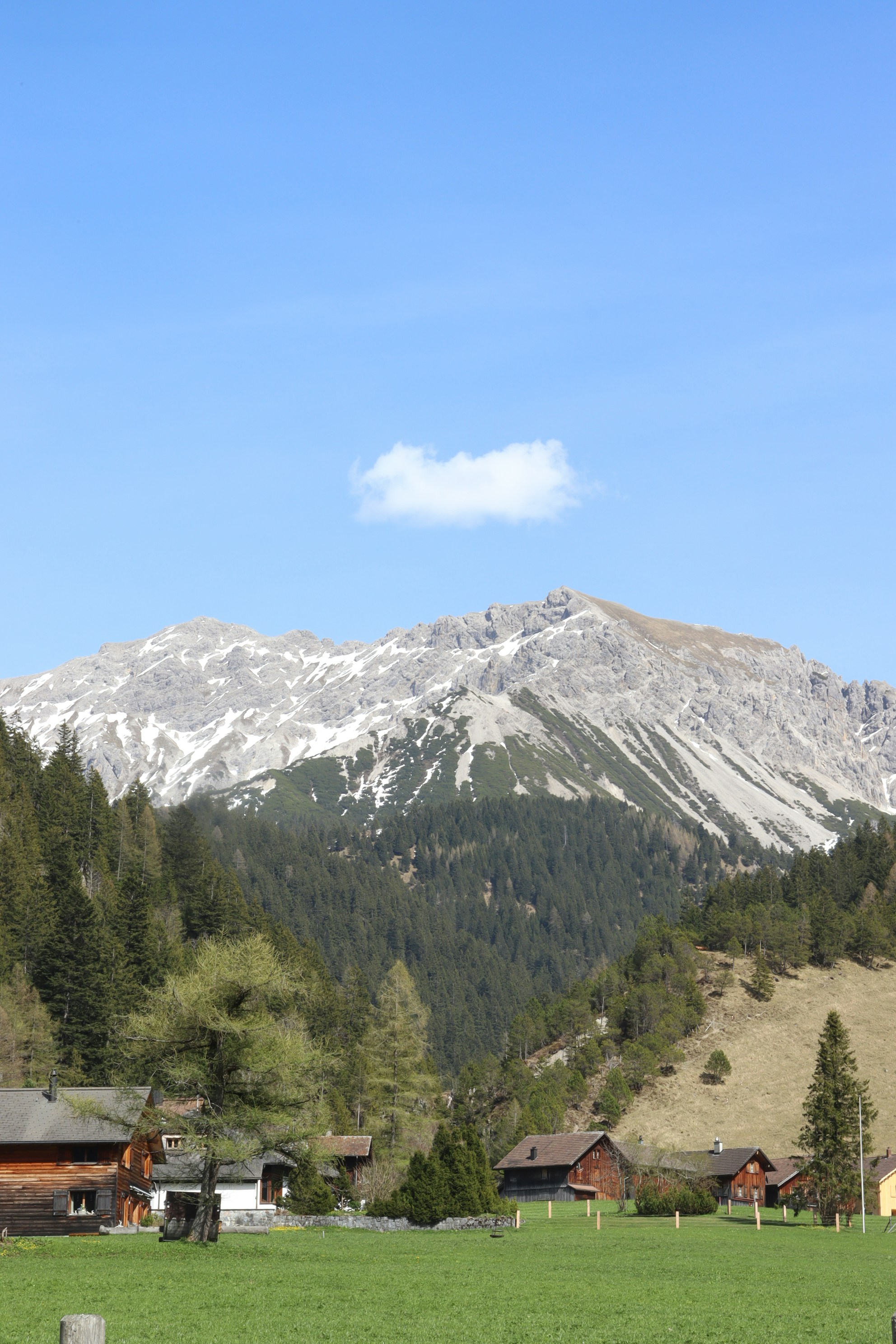 Cascade of green valley village at base of snow-capped peaks under a lone cloud