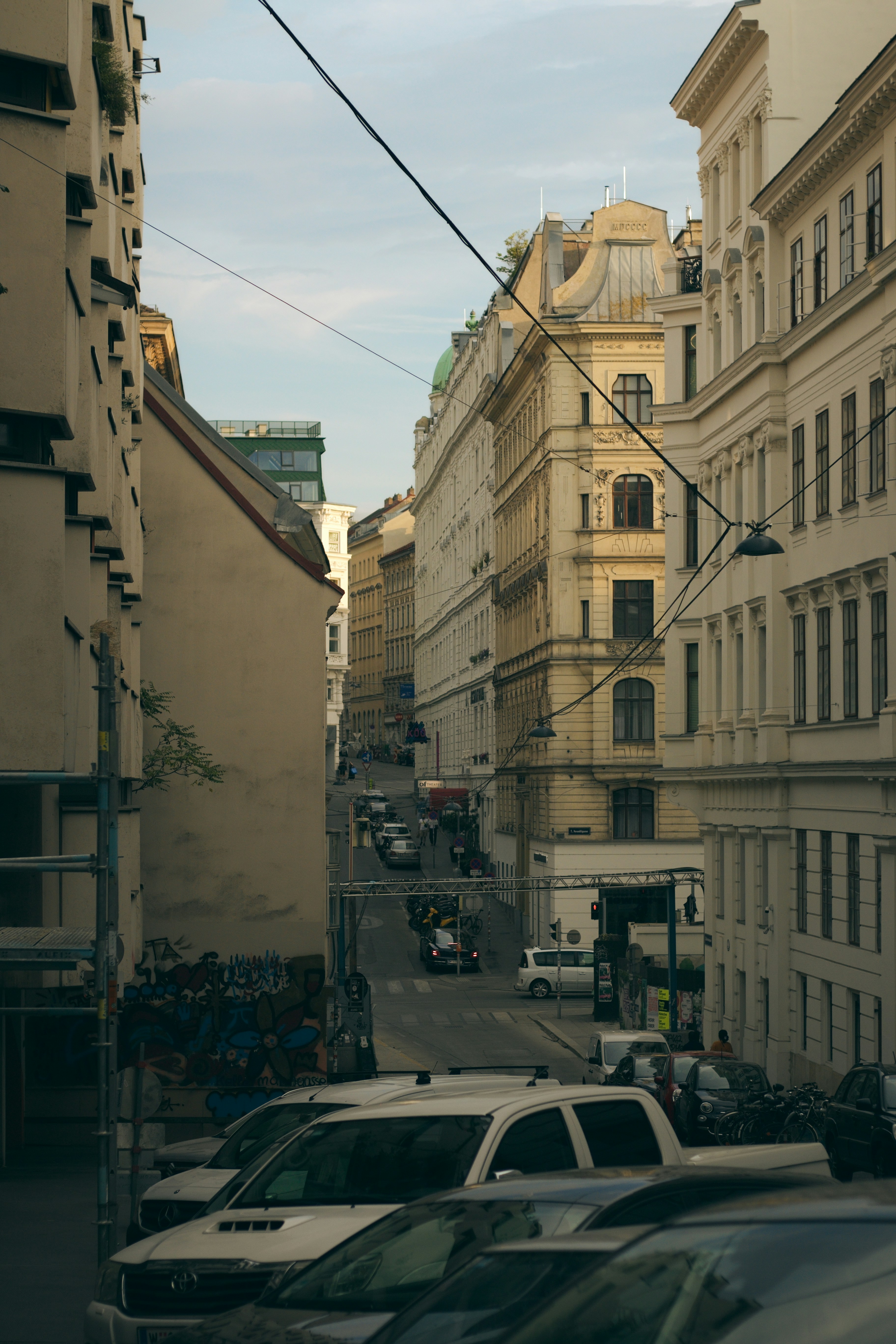 A narrow urban street lined with classical buildings, cars parked along both sides, and overhead cables crossing toward a distant, sunlit vanishing point.