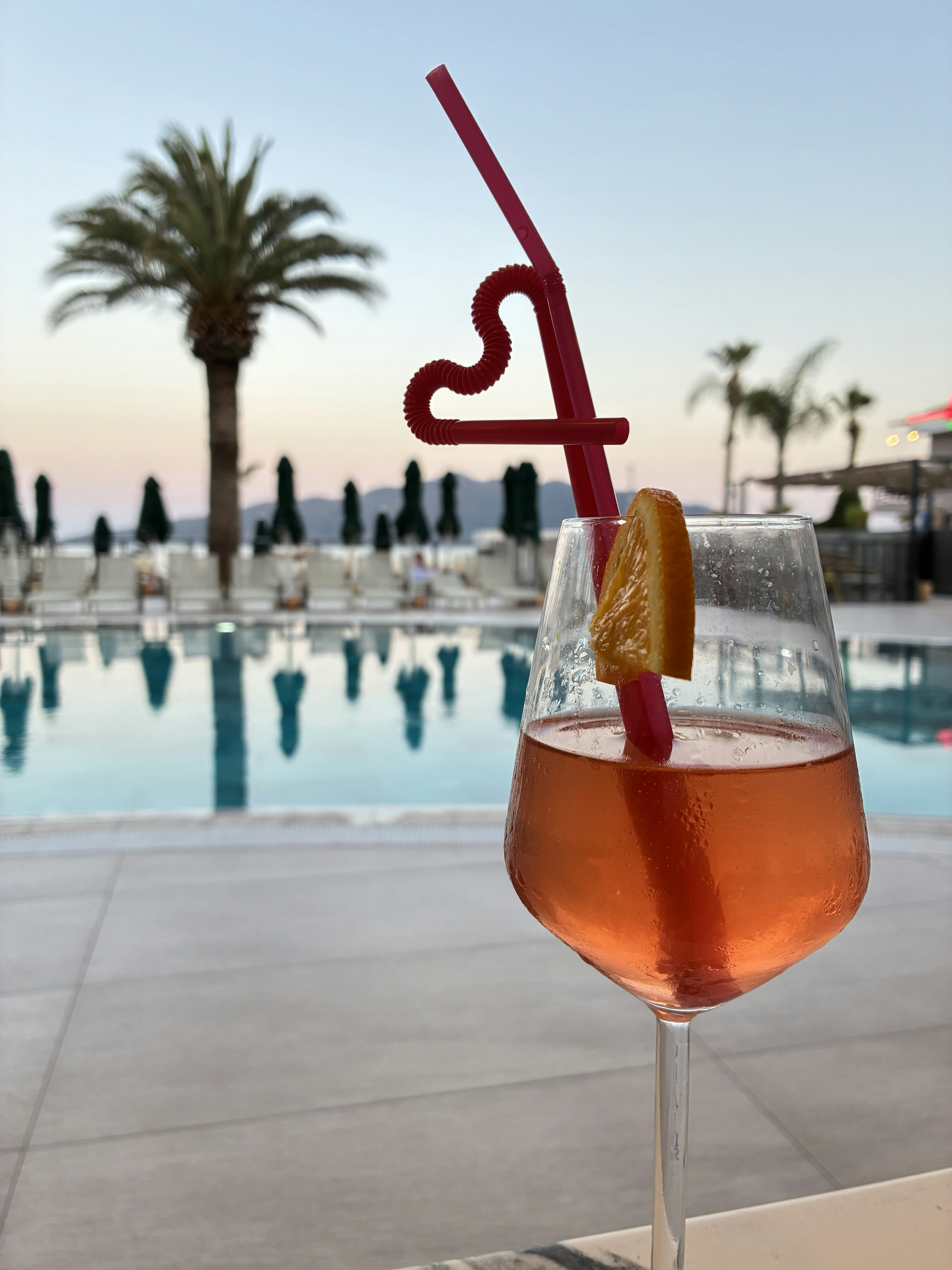 A bright orange cocktail with a decorative straw and orange wedge sits at the pool edge, with palm trees and a calm pool reflecting the horizon in the background.