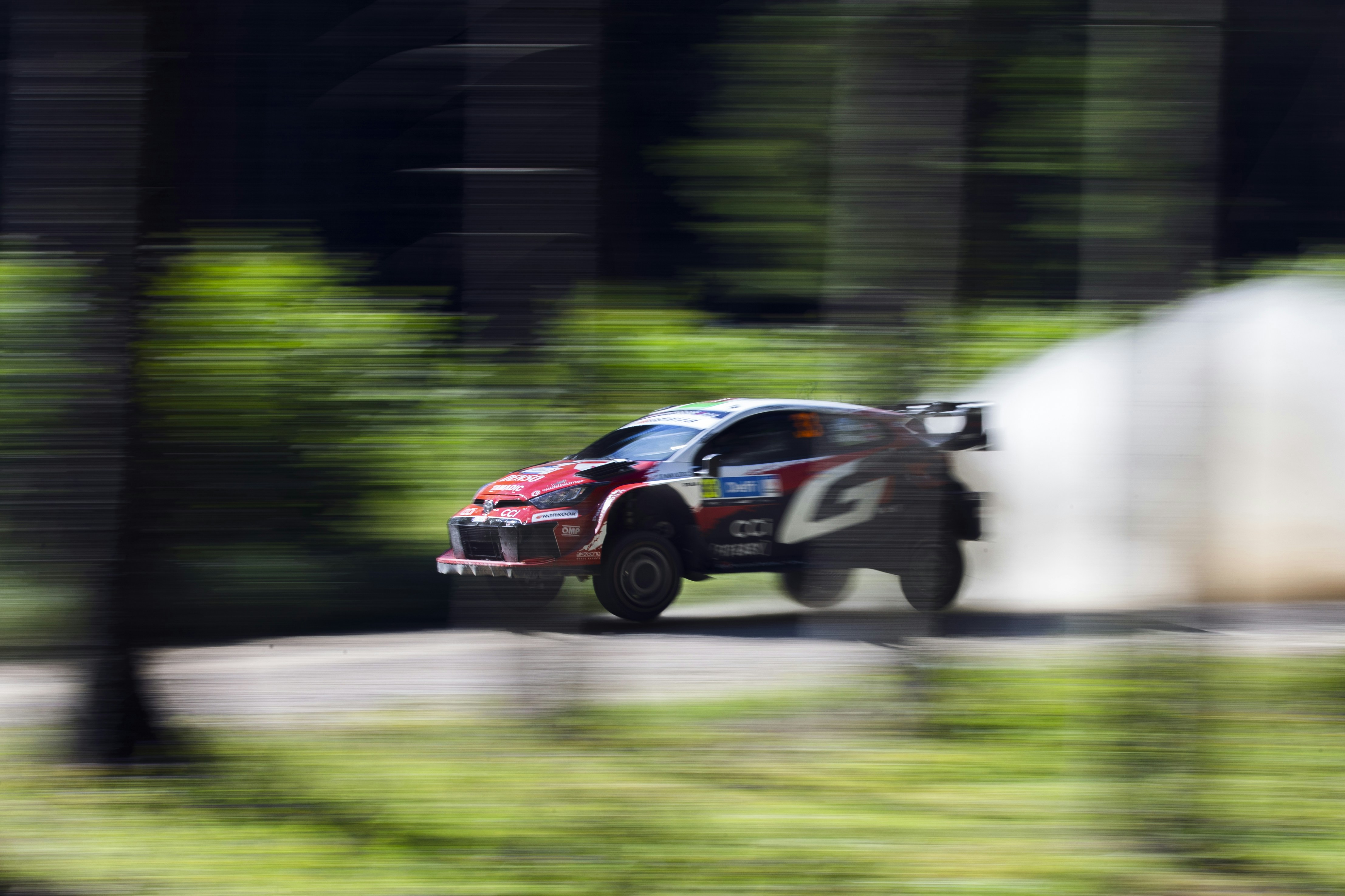 Red rally car blasting along a track with motion blur suggesting high speed and a forested backdrop.