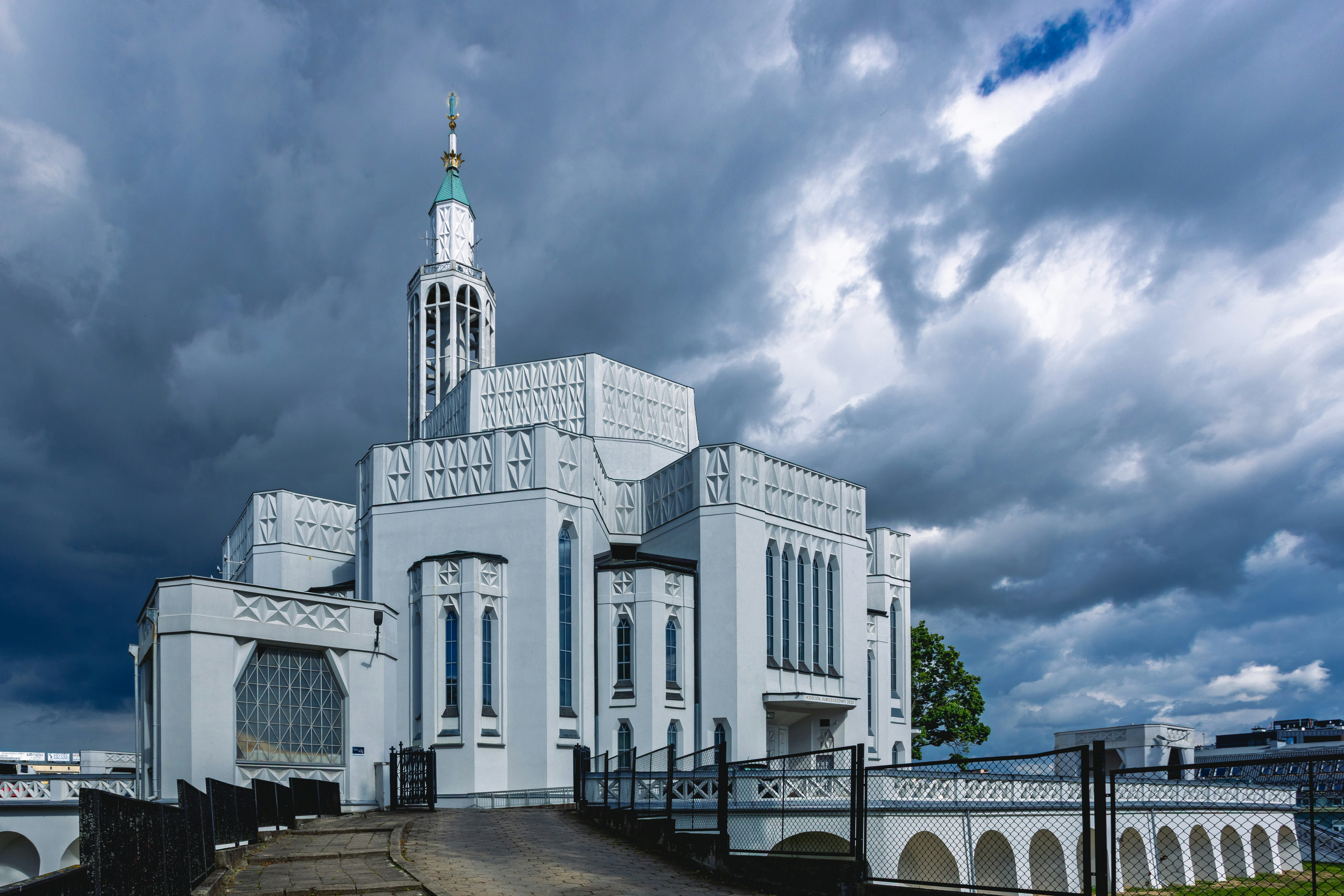 A white, art-deco style building sits on a riverside promenade beneath dramatic storm clouds, with a small green tree behind and railings along the walkway.