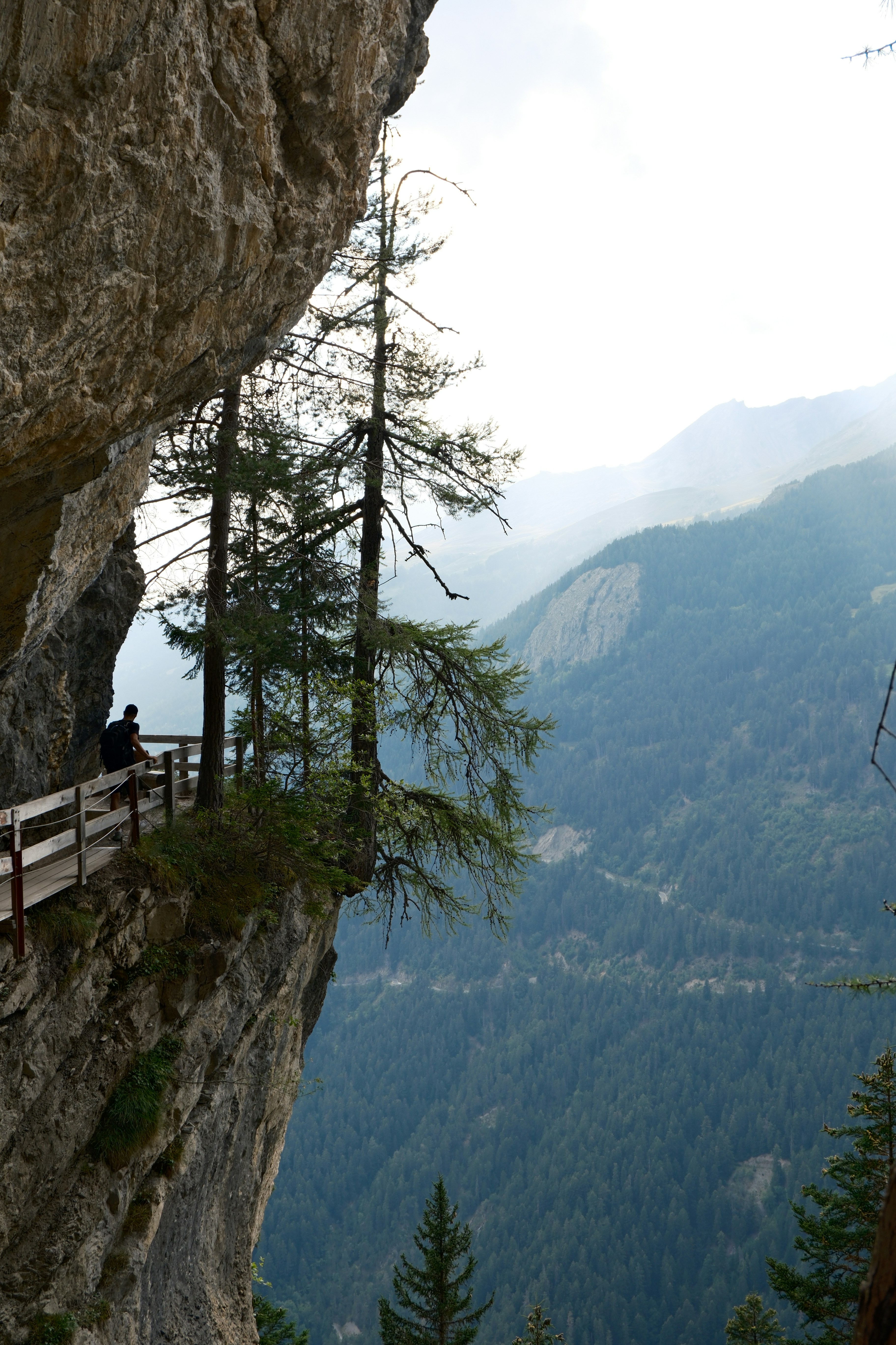 A narrow wooden walkway hugs a steep cliff, with hikers pausing near the railing as forested valleys unfold below.