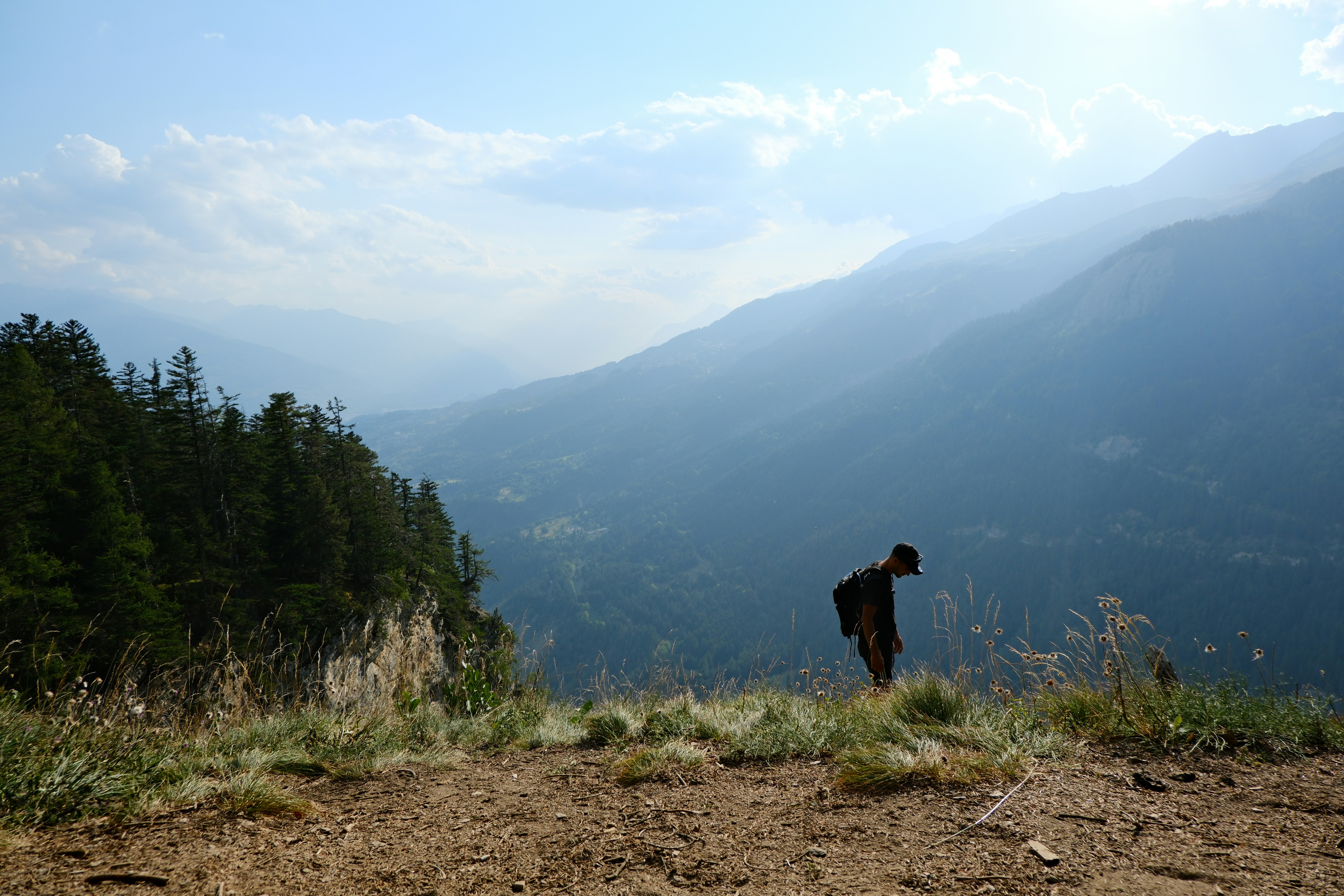 An alpine hiker pauses on the edge of a mountain ridge, surrounded by endless forests and layered peaks fading into the distance. Soft rays of sunlight break through the clouds, creating a peaceful atmosphere that captures the freedom, solitude, and beauty of exploring the high mountains. A timeless landscape inviting adventure and quiet reflection.