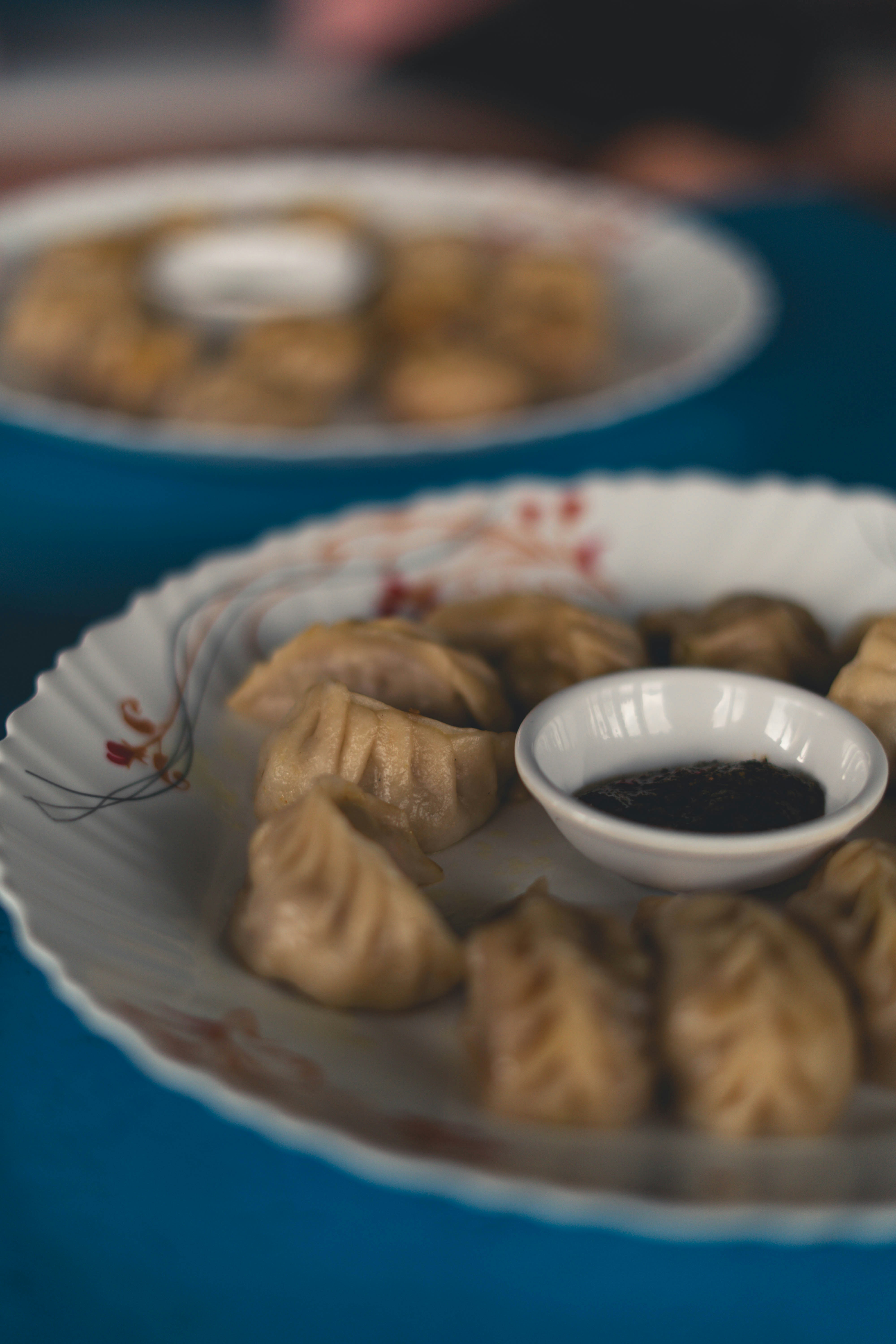 Close-up of a plate of dumplings with a small bowl of dark sauce, set on a blue table. Clear focus on the front dumplings while other dish in background is blurred; note logo/plate design and texture of dough.