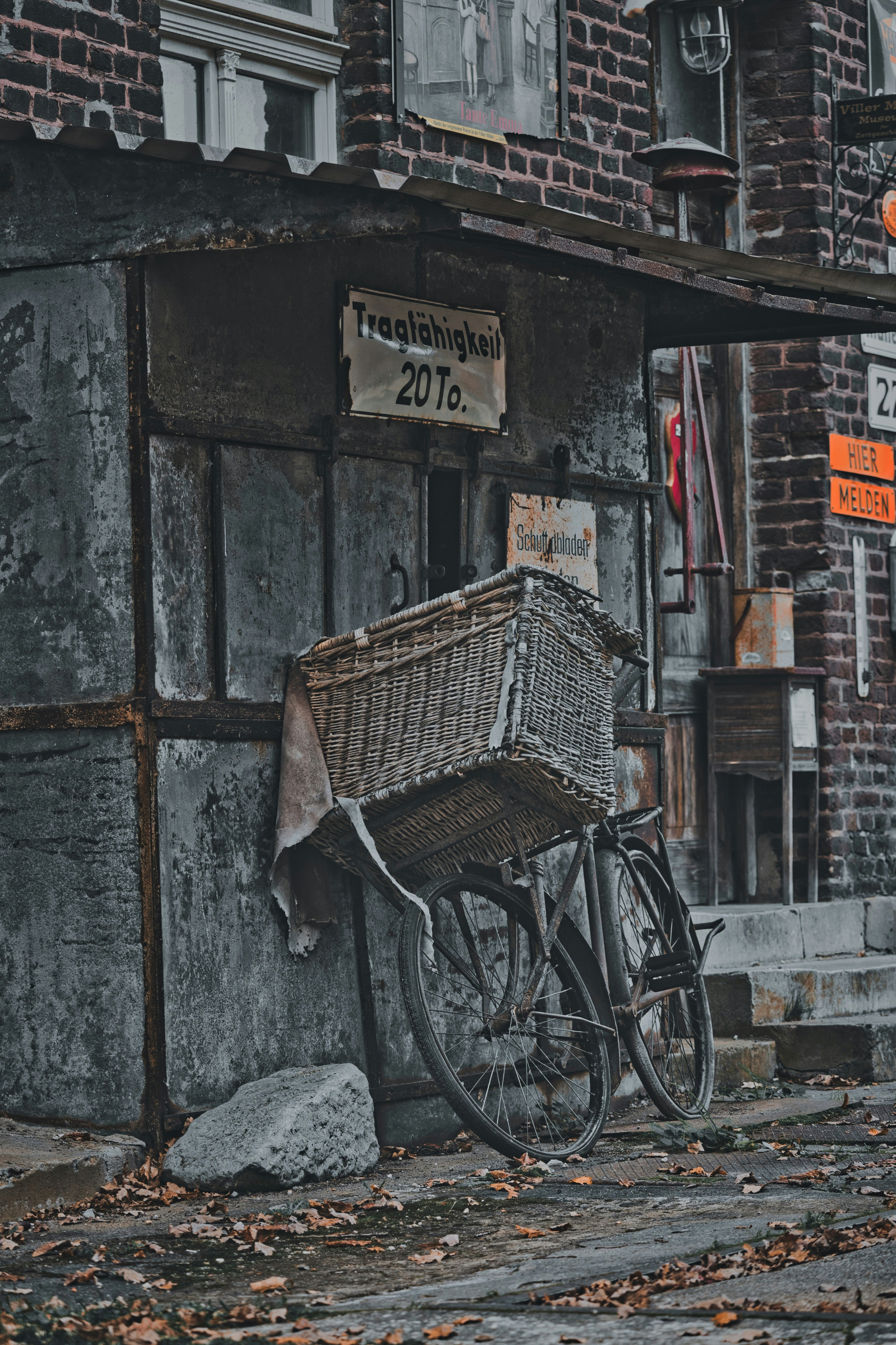 Weathered bicycle with a large wicker basket leans against a deteriorated storefront on a gritty urban street; autumn leaves litter the wet pavement near a brick wall.