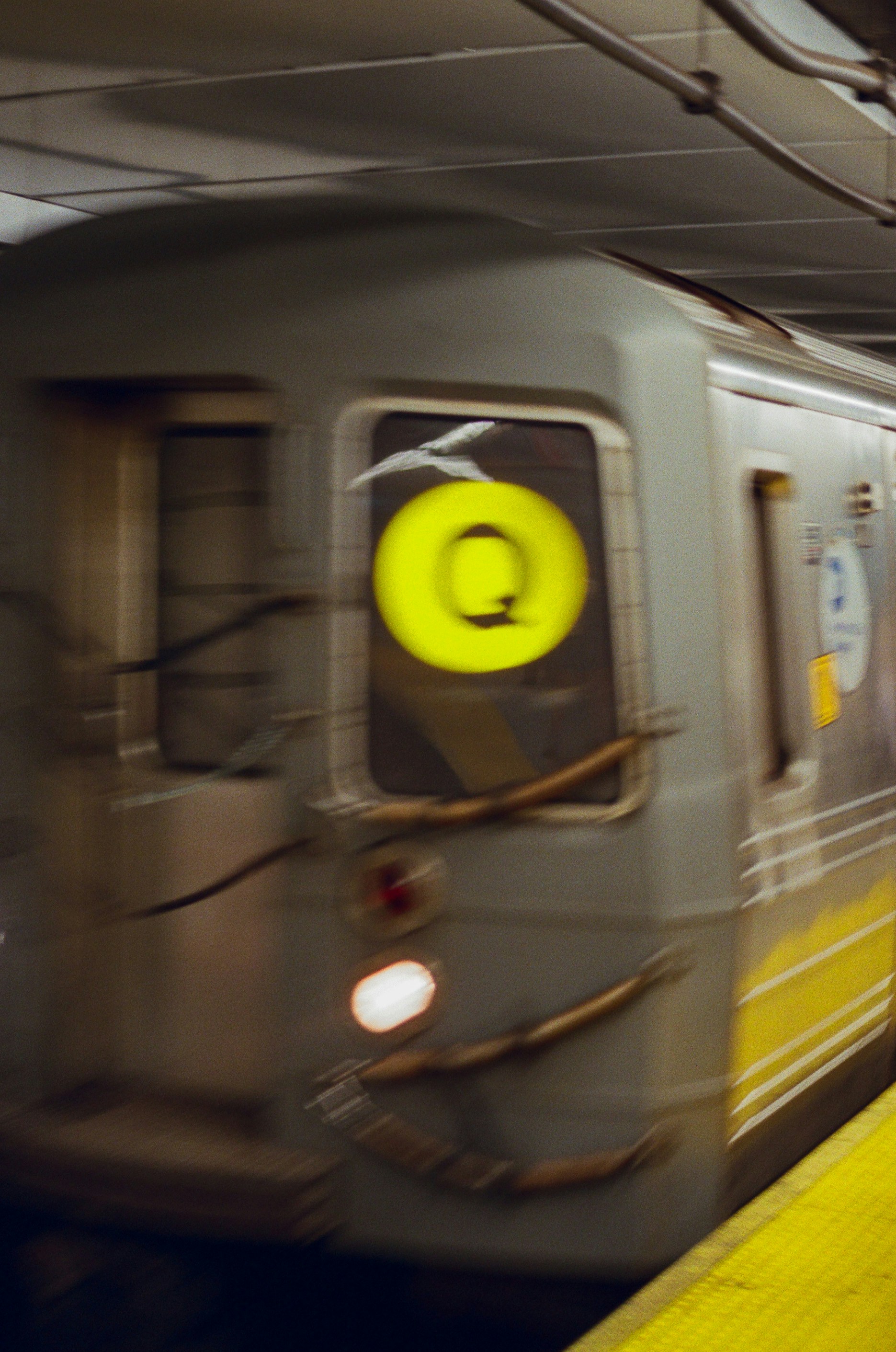 A subway train rushing through a station, captured with motion blur on film and illuminated by a vivid yellow route sign.