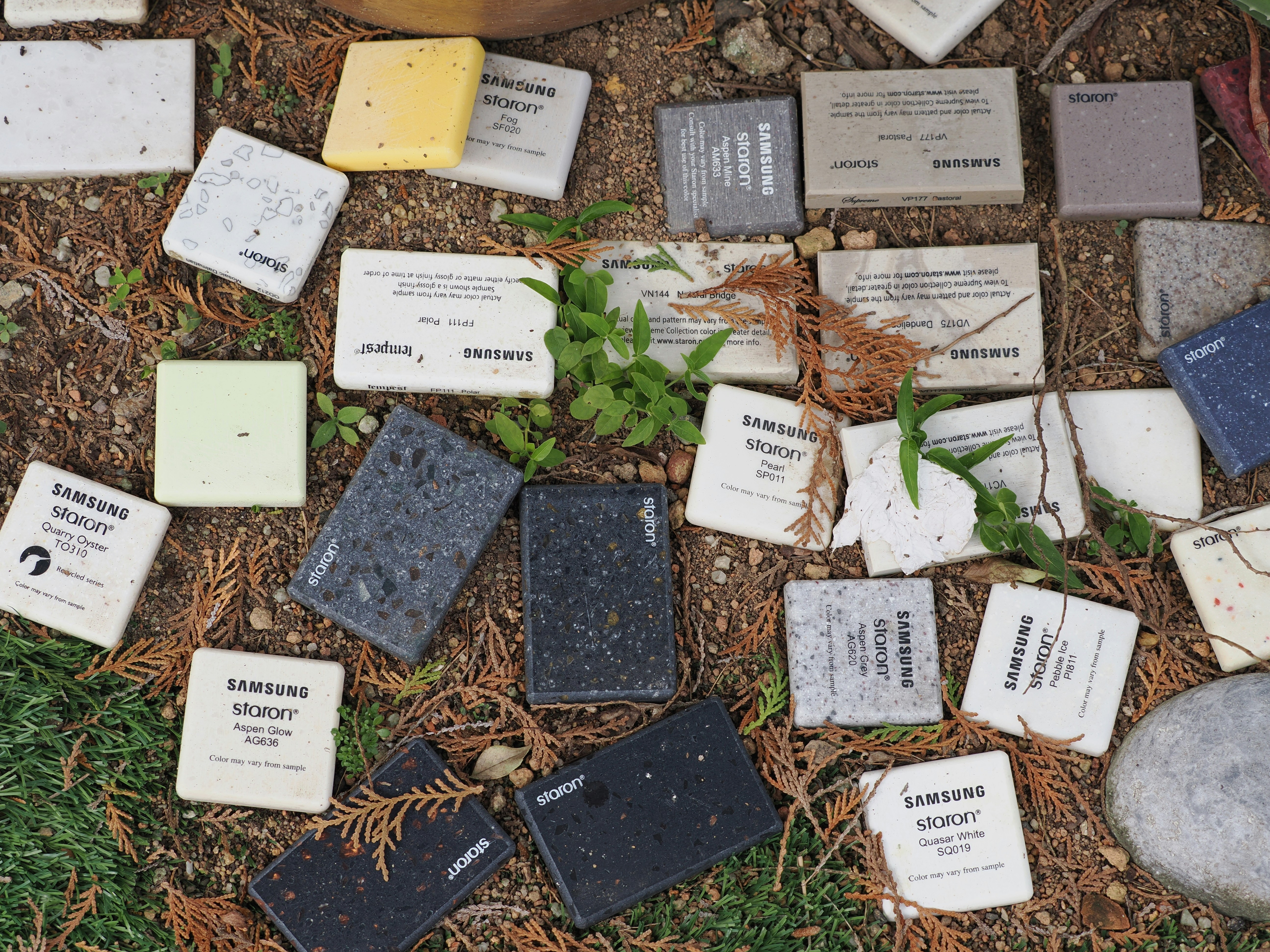 A ground-level mosaic of scattered Samsung memory tiles and pavers, with greenery peeking through; the arrangement forms a textured pattern on soil and grass.