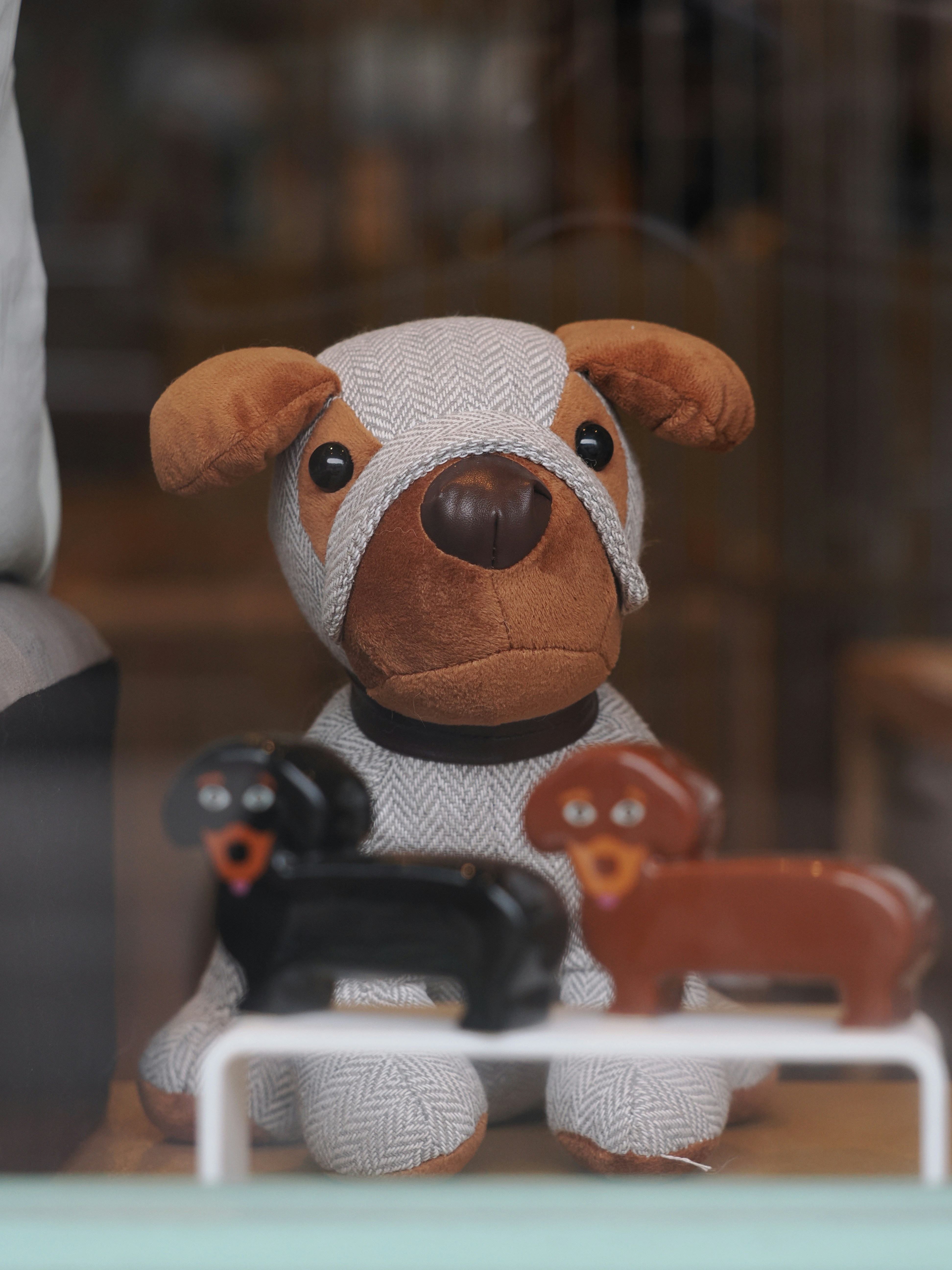 A plush dog wearing a herringbone hoodie sits behind a small display of toy horses, viewed through a storefront window.