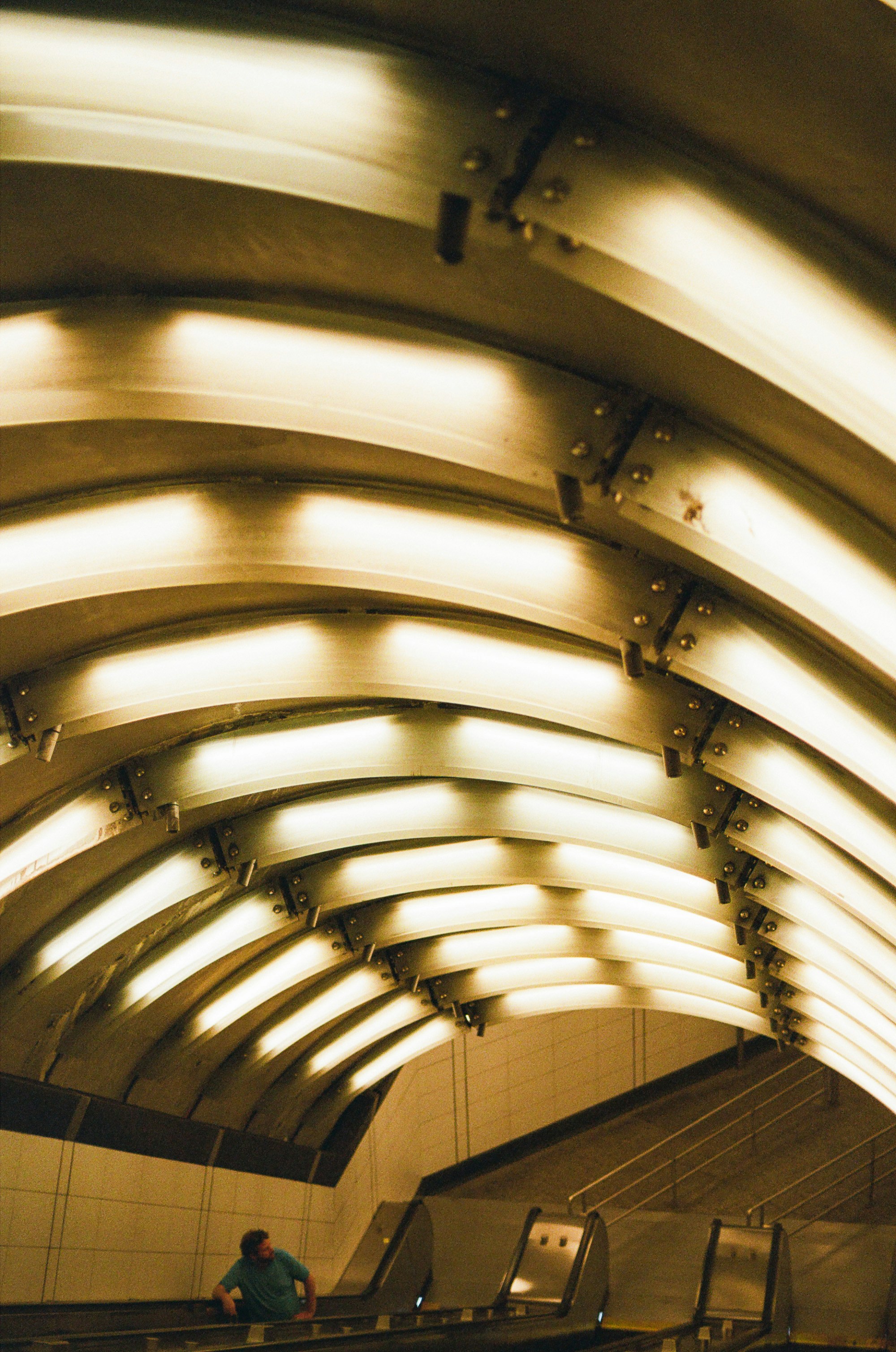 A lone passenger riding an escalator beneath curved fluorescent lights, creating a quiet and cinematic underground scene.