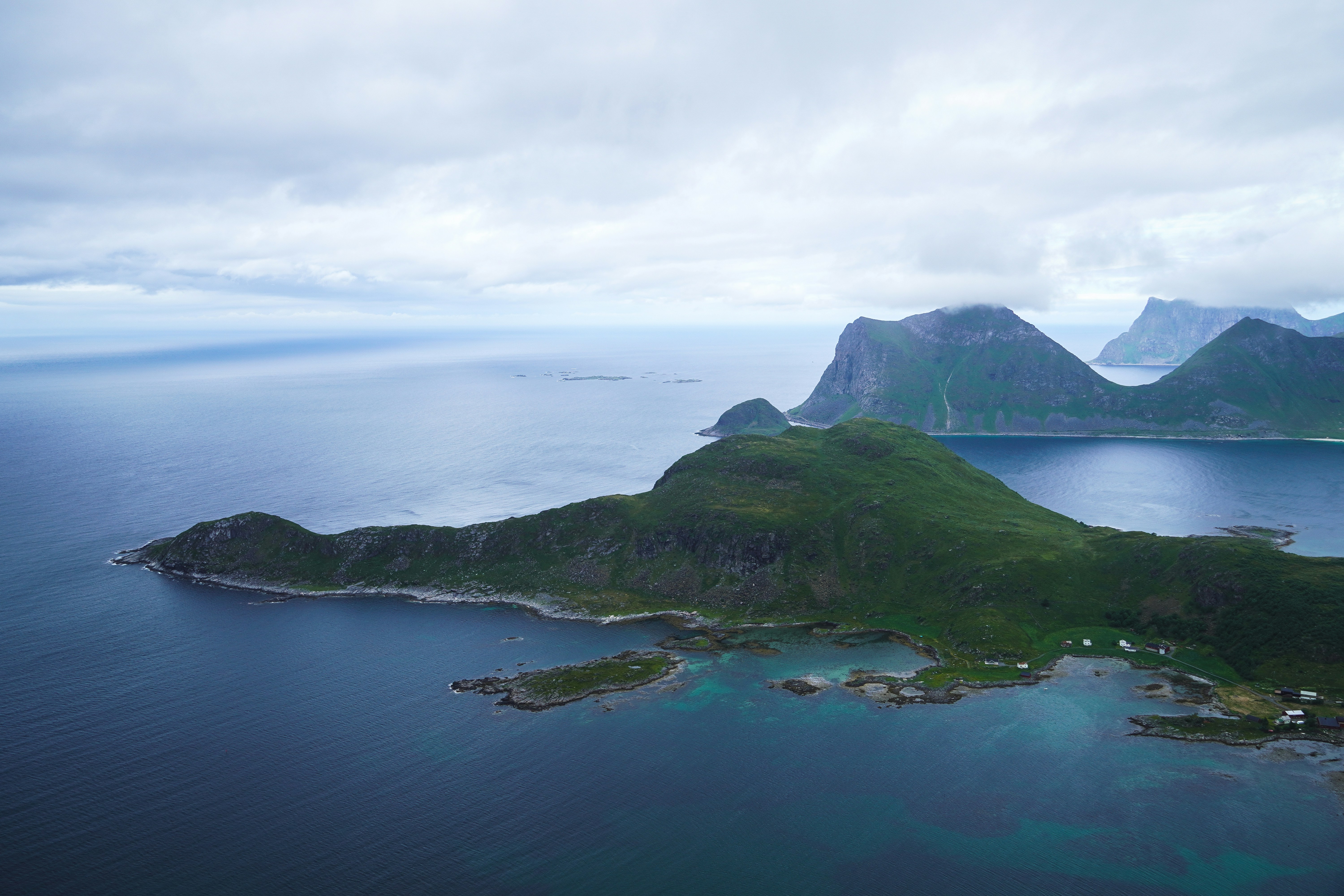 Coastal island arch extending into calm blue sea with turquoise shallows and distant hills under a cloudy sky.
