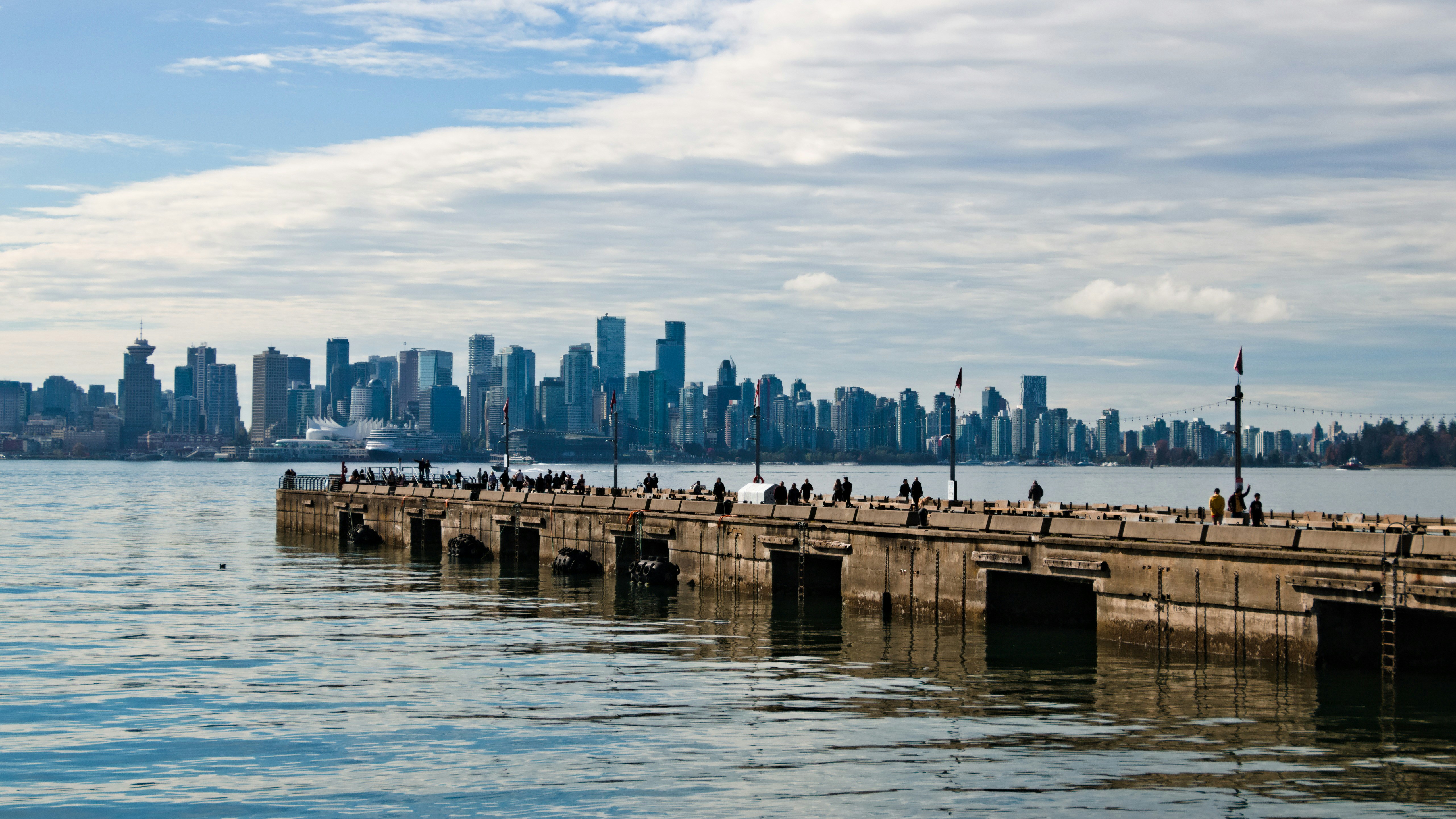 Wide pier extends into quiet water with a distant city skyline under a partly cloudy sky; figures line the jetty, their silhouettes contrasting with the blue water.