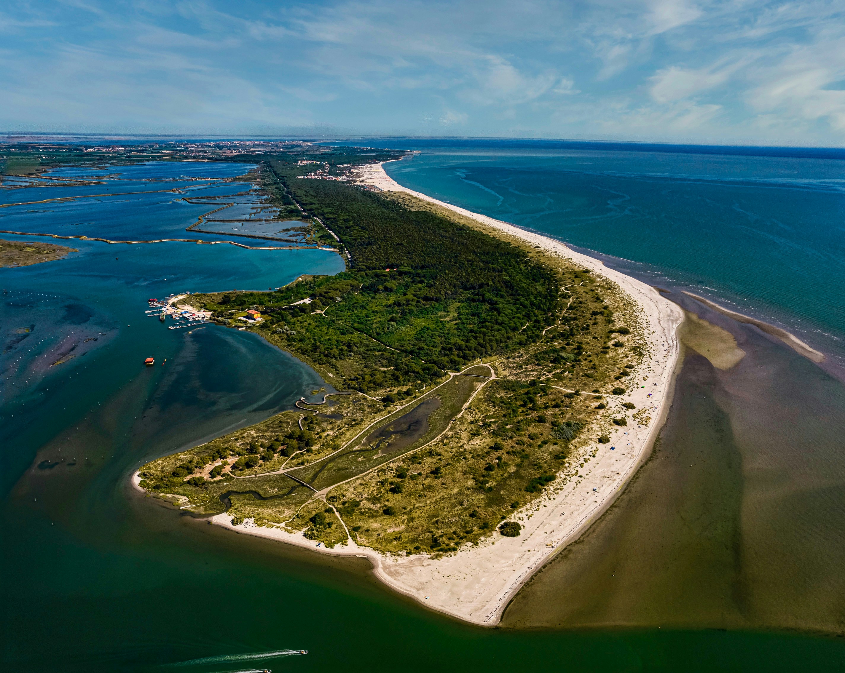 Airborne view of a sandy, crescent-shaped sandbar extending into a blue-green lagoon, with lush vegetation covering the interior and calm tidal channels on the sides.