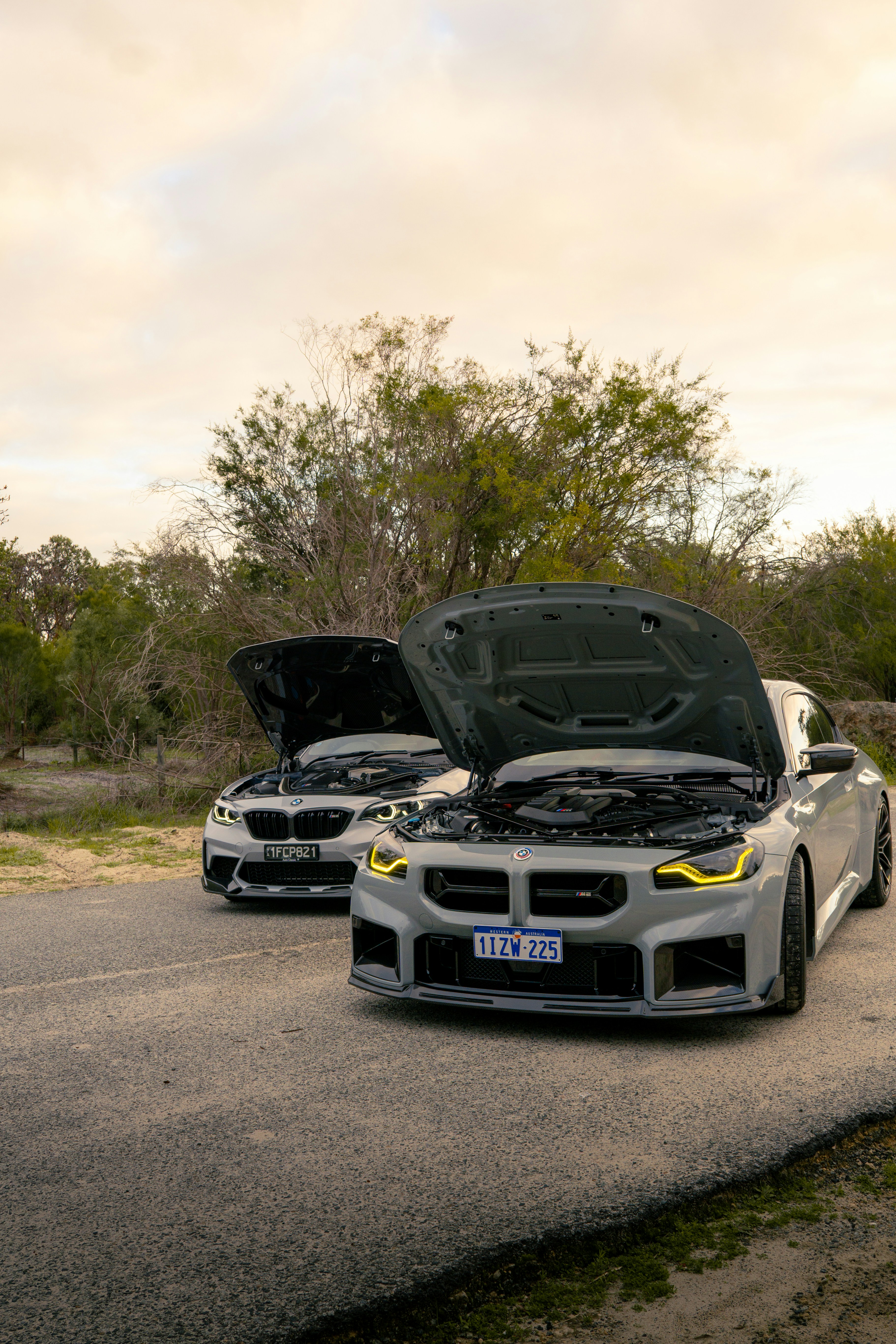 Two high-performance cars with hoods open sit in a parking area, revealing their engines against a backdrop of trees at dusk.