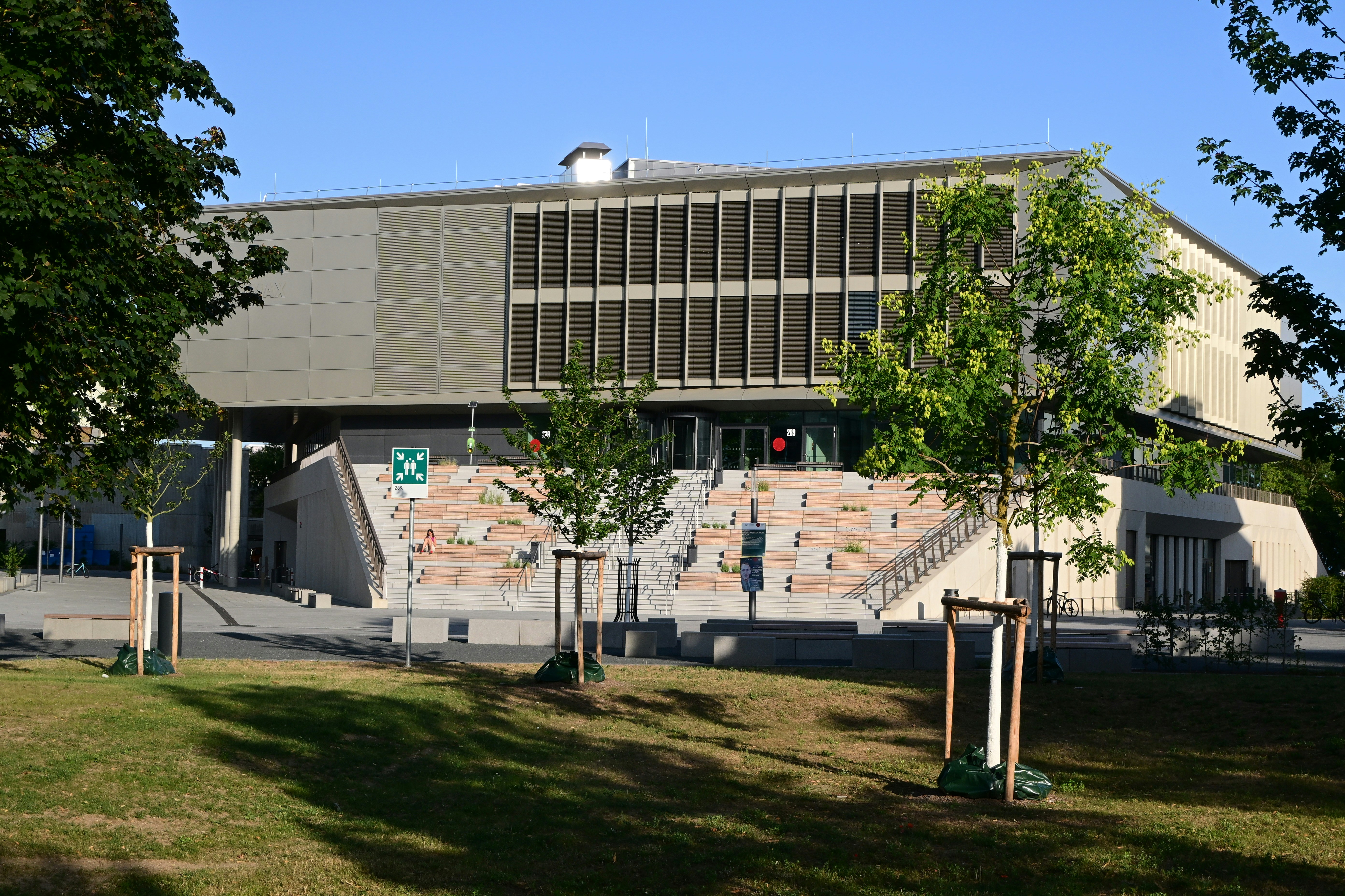 Sunlit modern building with stairs and glass facade sits behind a grassy park with young trees; a quiet, open campus scene with strong shadows and a clear blue sky.