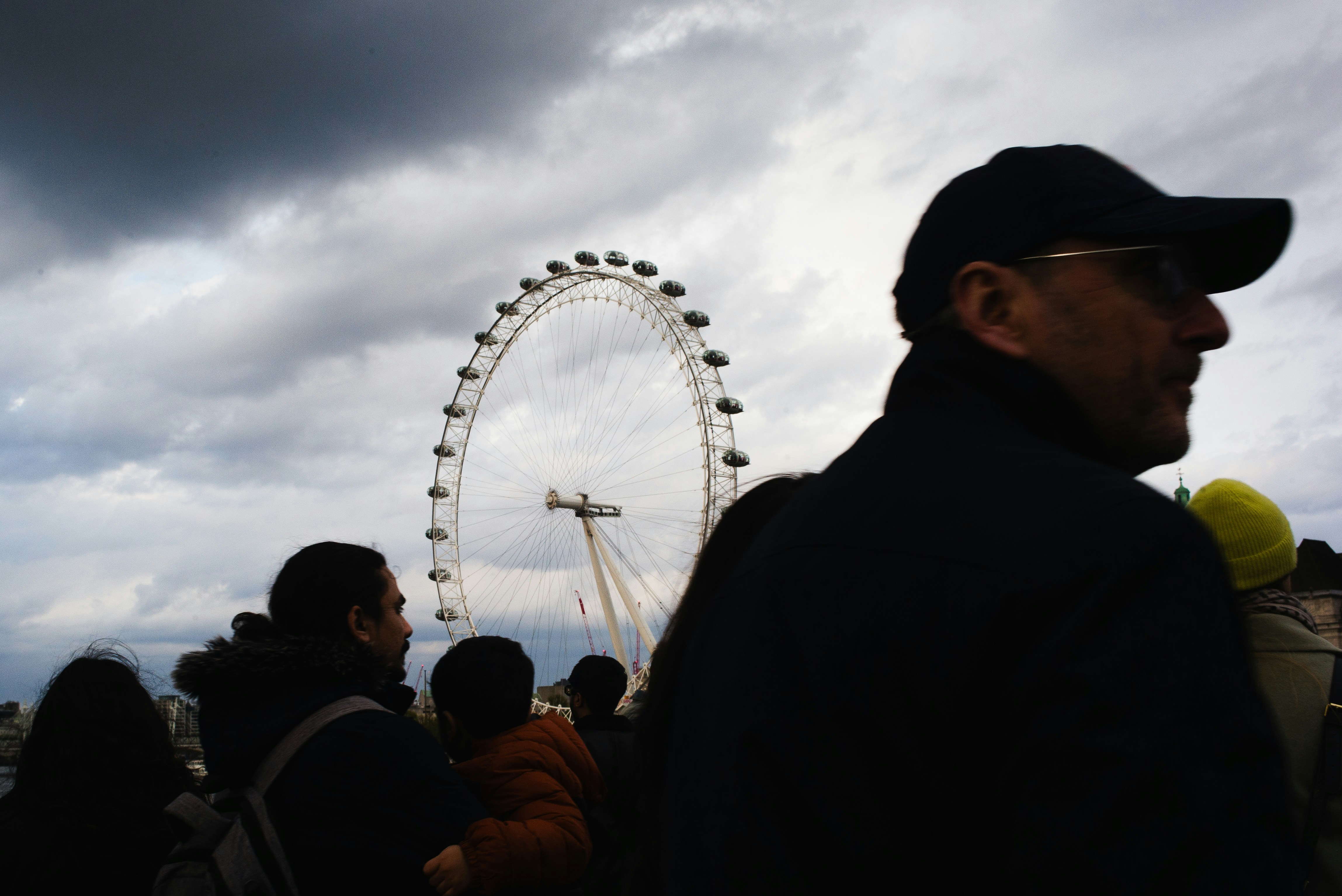 A crowded urban scene with silhouettes of people in the foreground and a ferris wheel rising behind them, framed against a cloudy sky.