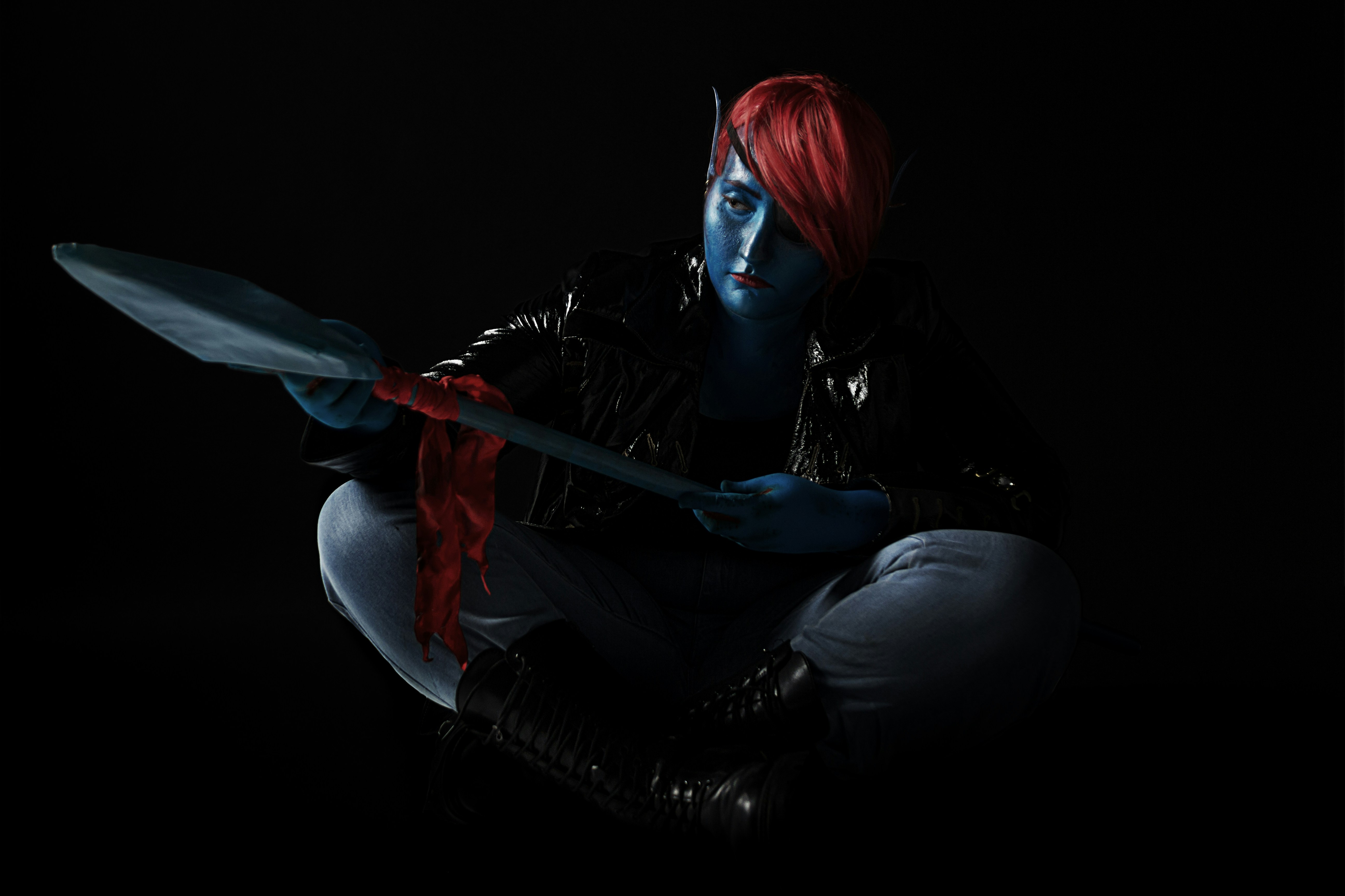 Blue-skinned character with red hair sits cross-legged in near-darkness, wielding a pale blade with a red cloth, emphasizing a dramatic, stylized portrait.