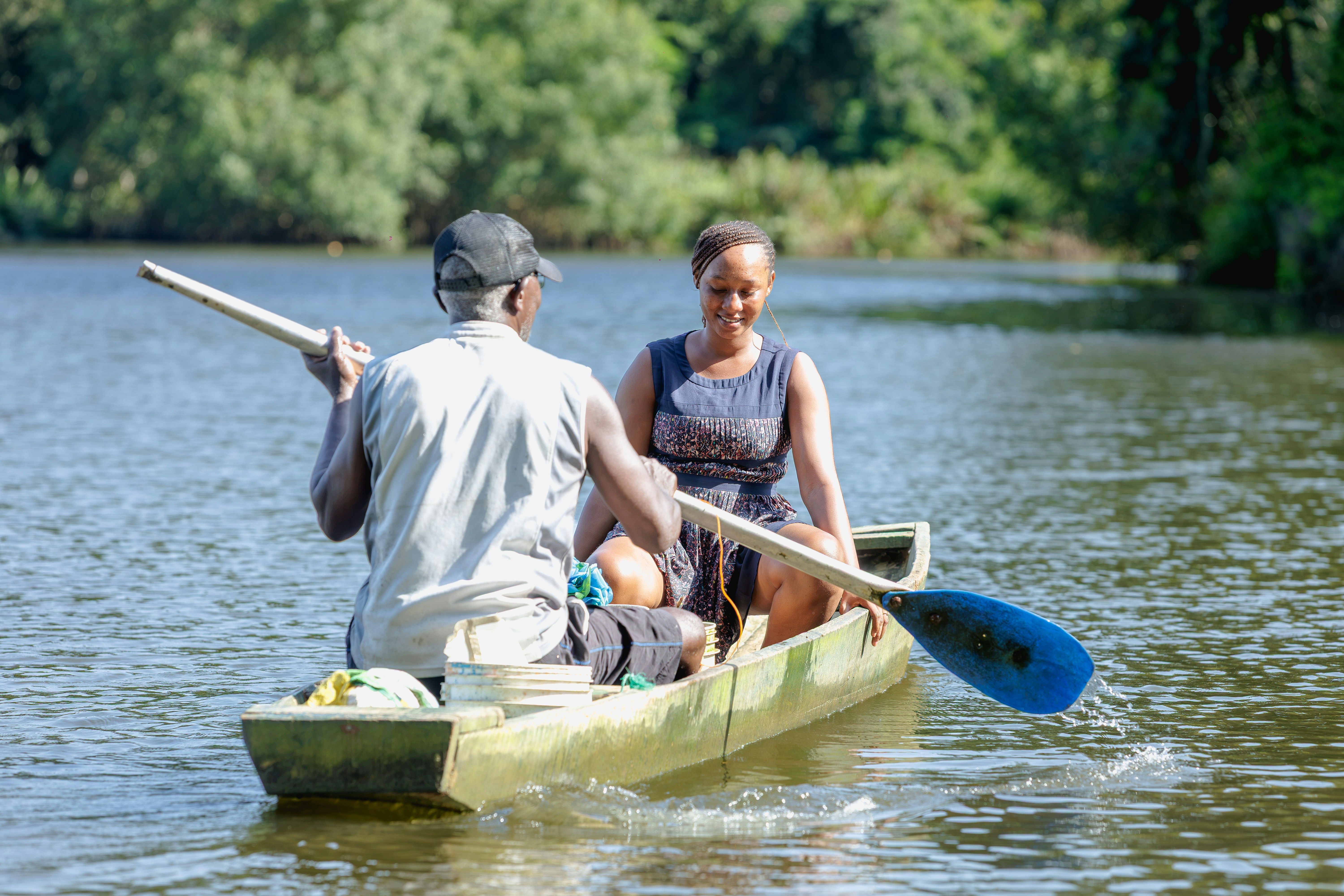 Two canoeists paddling a weathered boat along a sunlit river, with dense green foliage in the background.