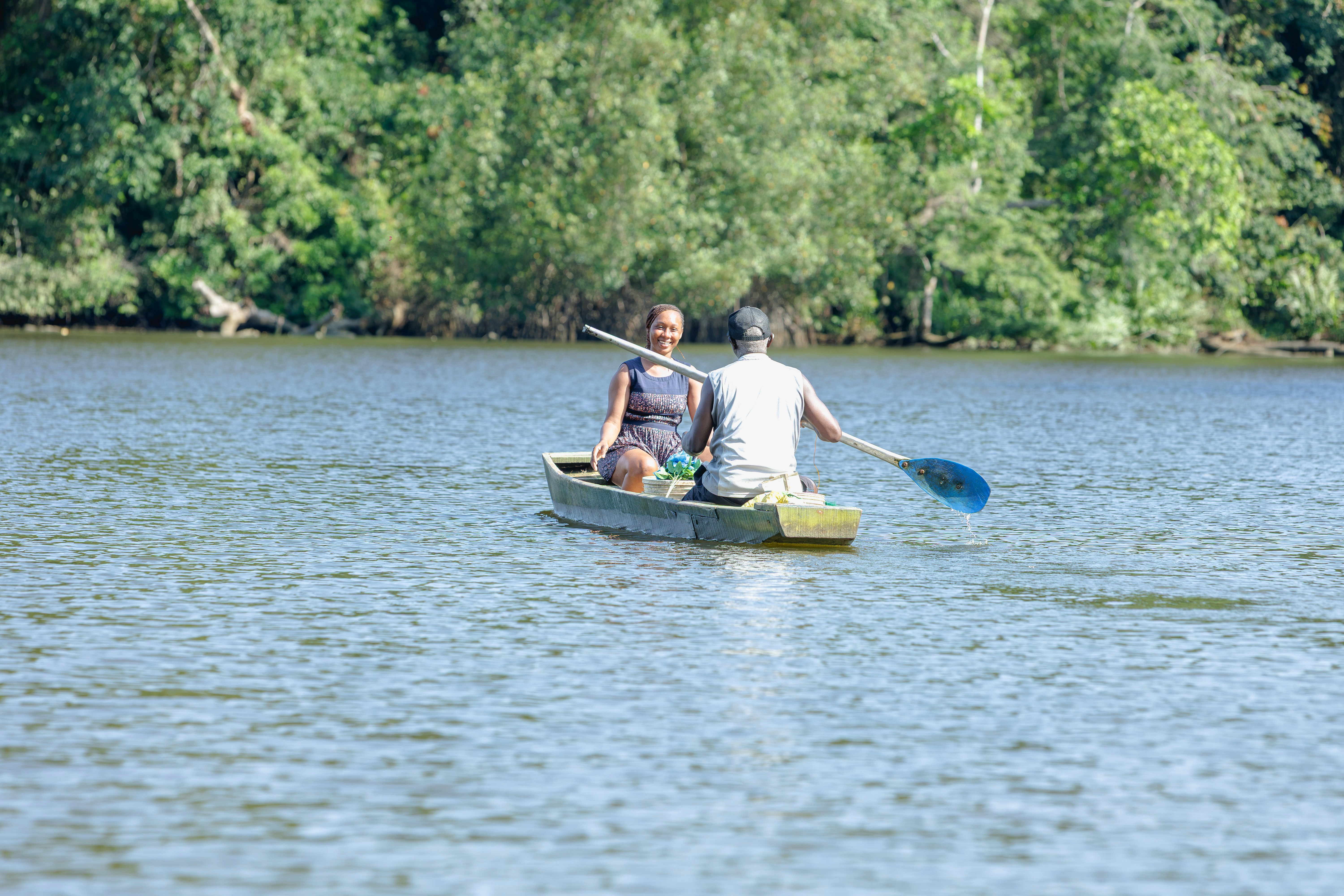 Two people paddle a small canoe on a calm lake with a tree-lined shore in the background.