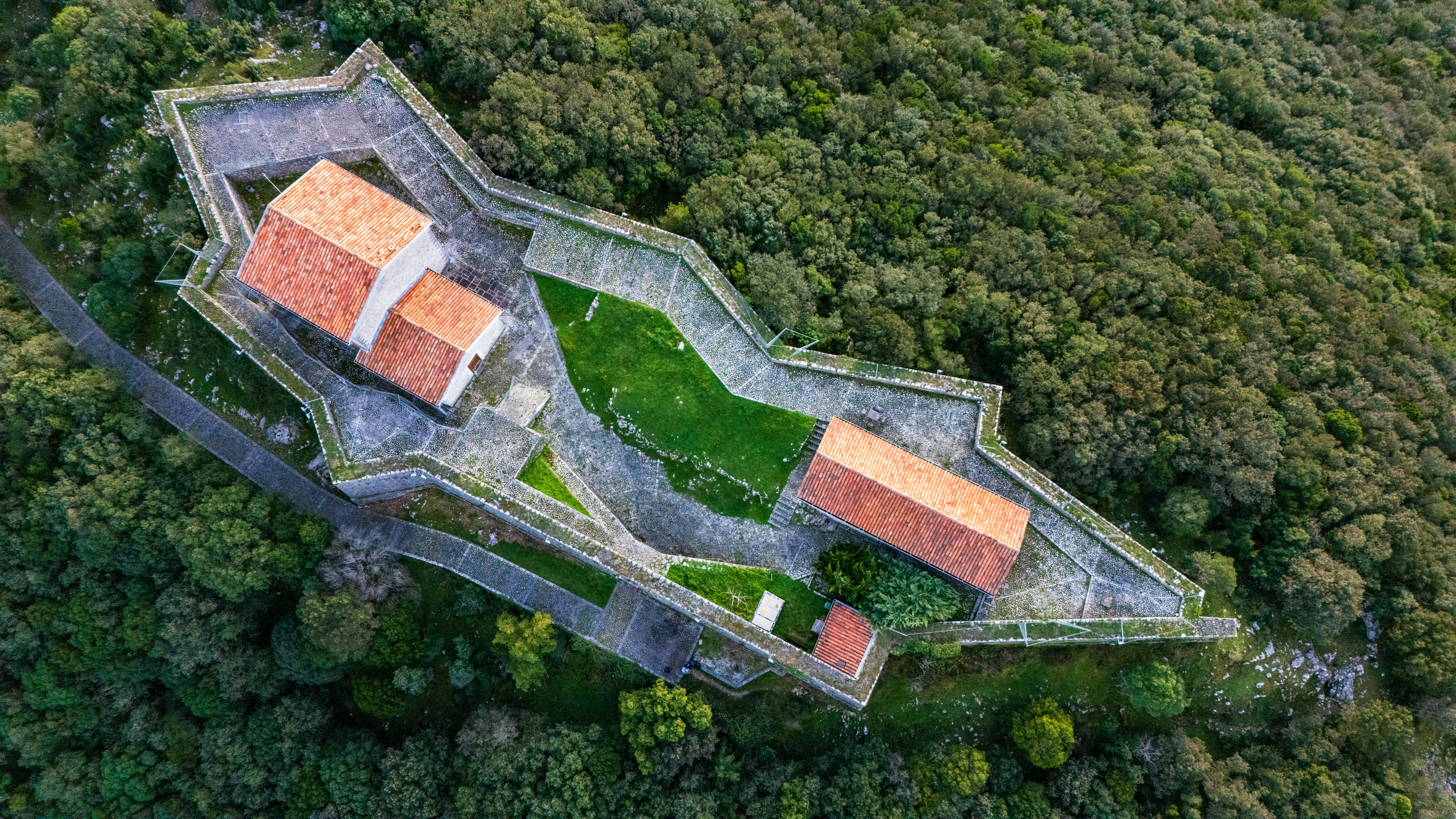 Aerial view of a stone fortress complex winding through dense forest, with red-tiled rooftops and green courtyards.