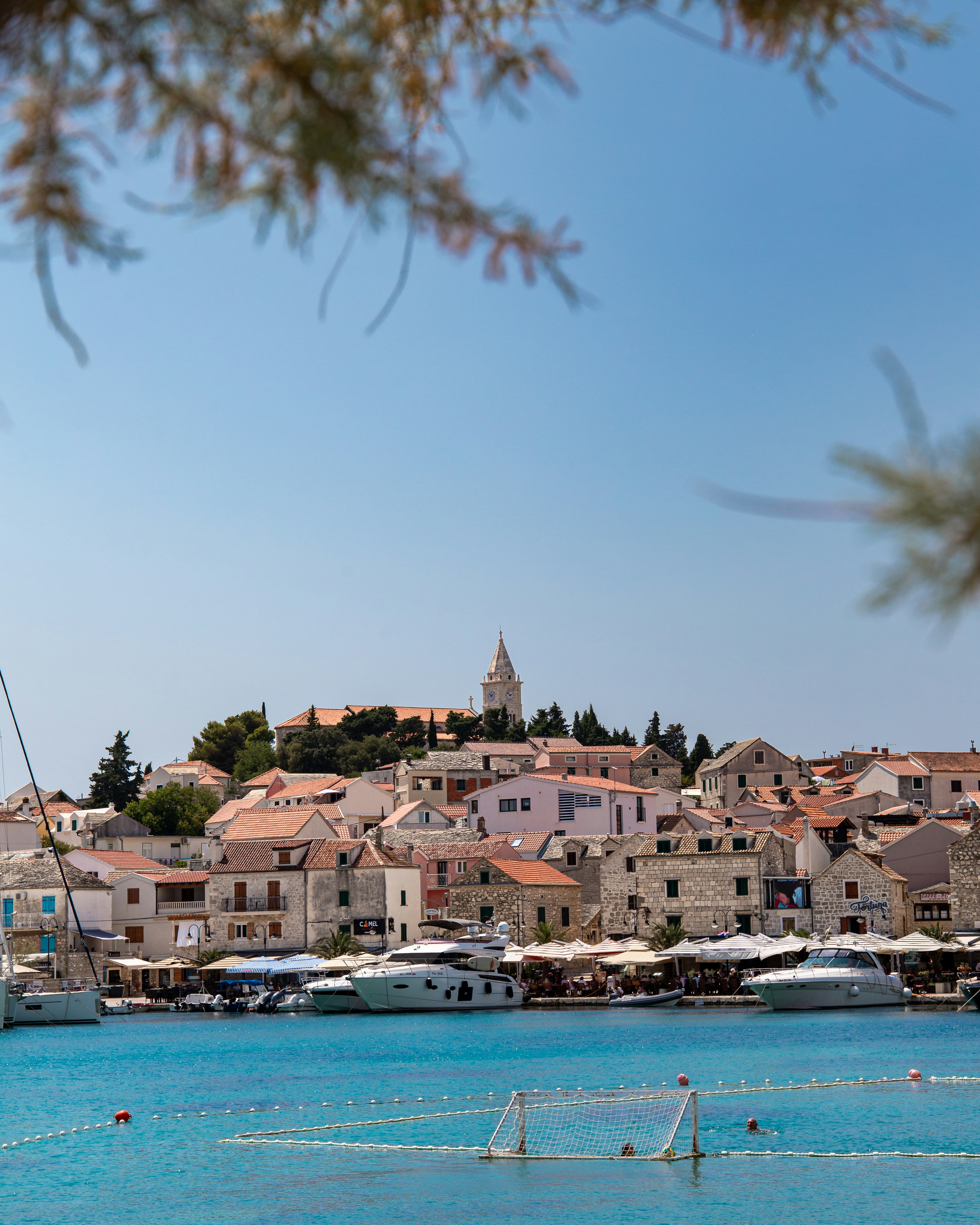 Coastal village with terracotta roofs along a turquoise harbor and distant church tower, boats docked at the quay, framed by tree branches at the top.