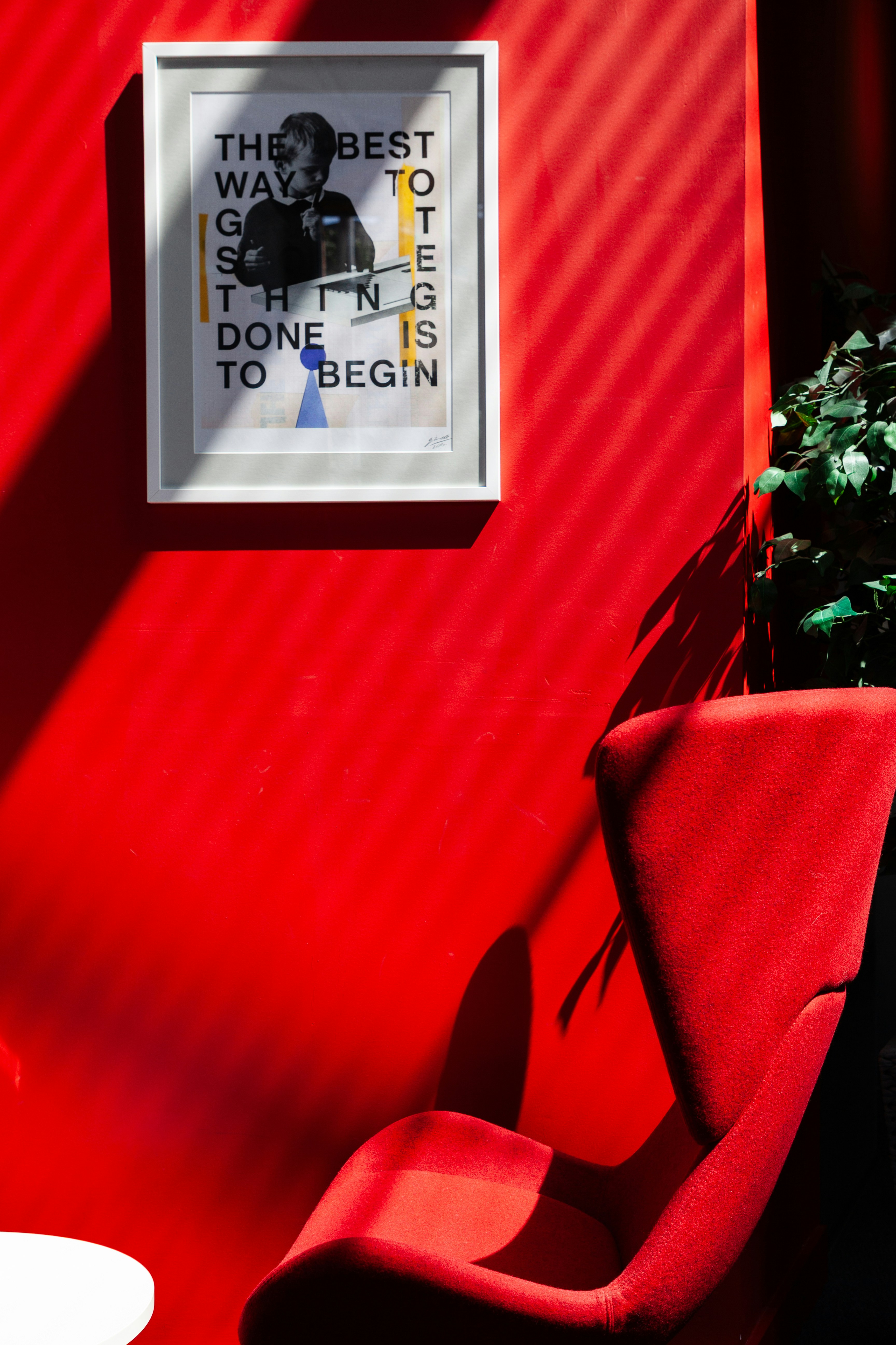 A framed black-and-white portrait hangs on a vivid red wall, with diagonal shadows crossing the scene and a red upholstered chair in the lower right. The setting emphasizes bold color contrast and modern decor.