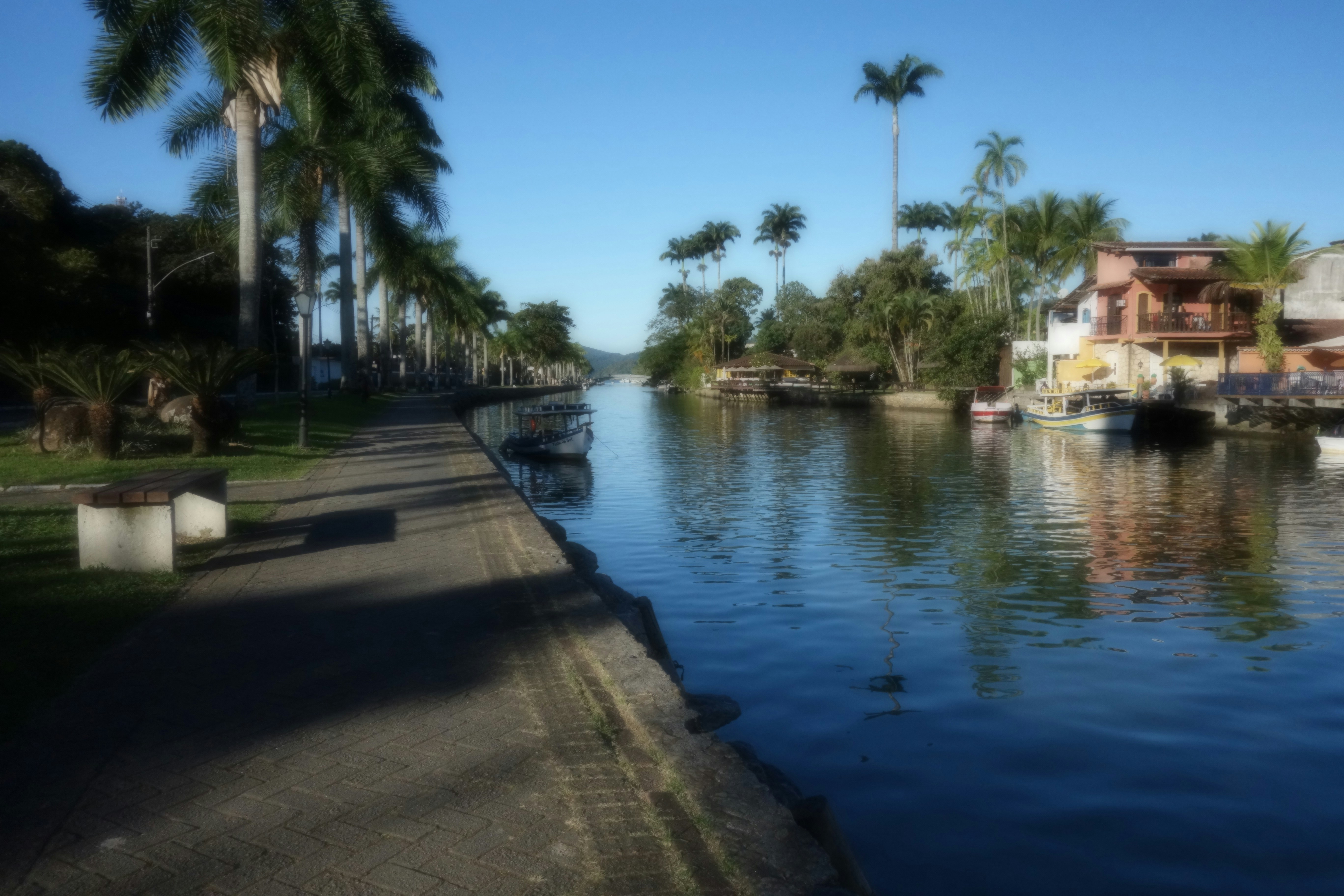 Sunlit canal scene along a paved promenade with calm water reflecting palms and buildings on the far bank; a tranquil riverside stroll captured along a linear waterfront.